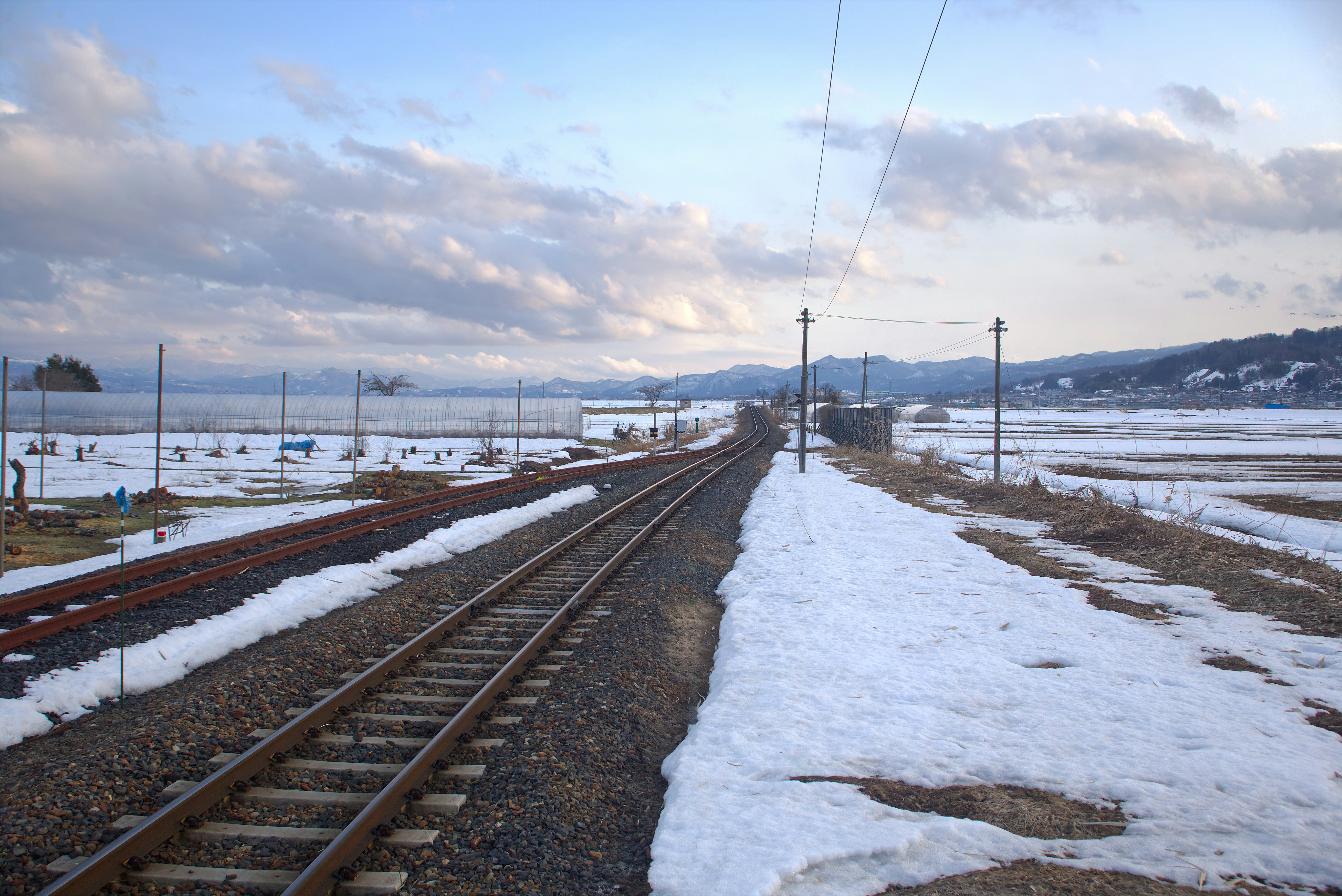 Railway tracks meandering through a snowy field with distant mountains under a cloudy sky.