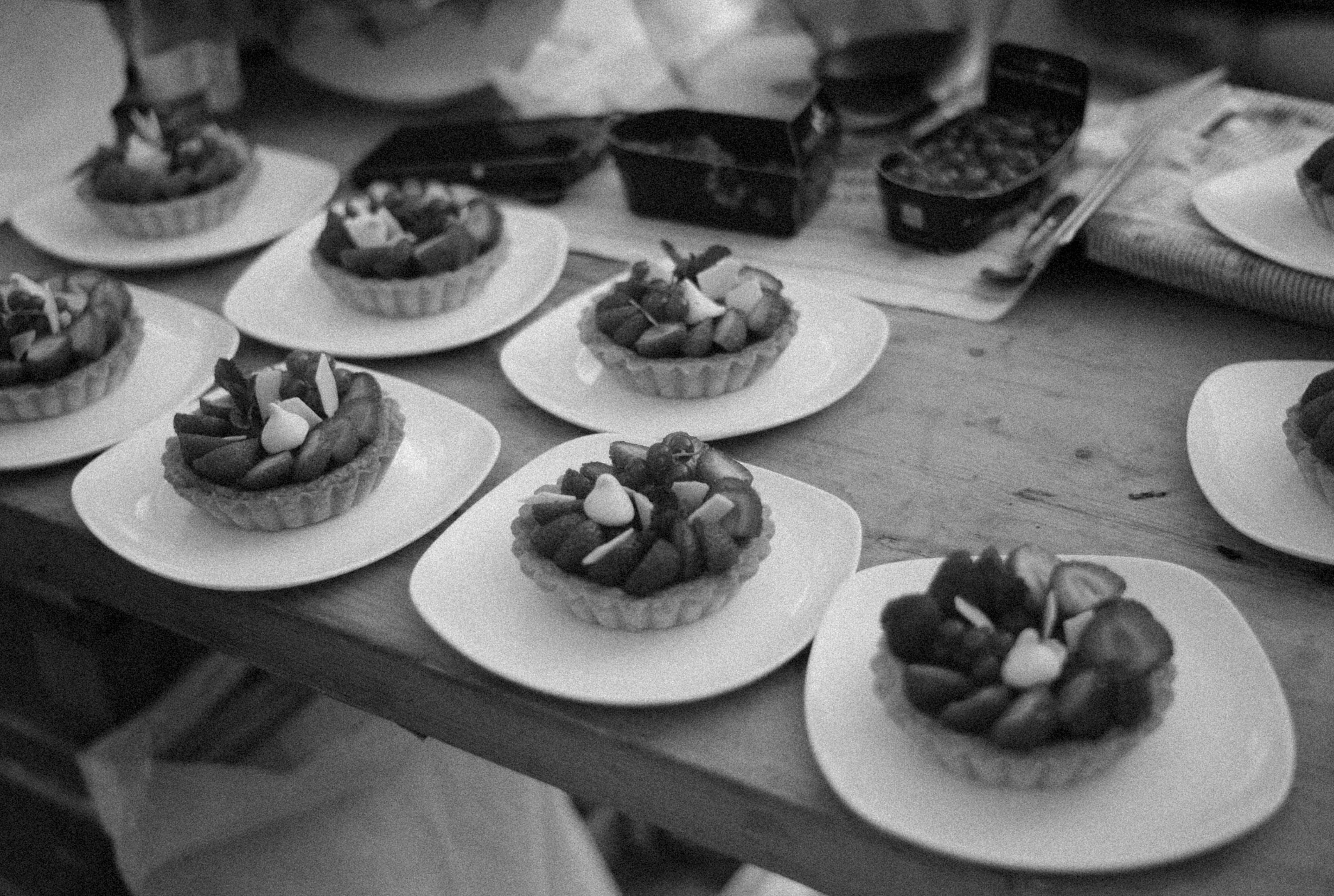 Monochrome photograph of small fruit tartlets arranged on white plates along a rustic wooden table.