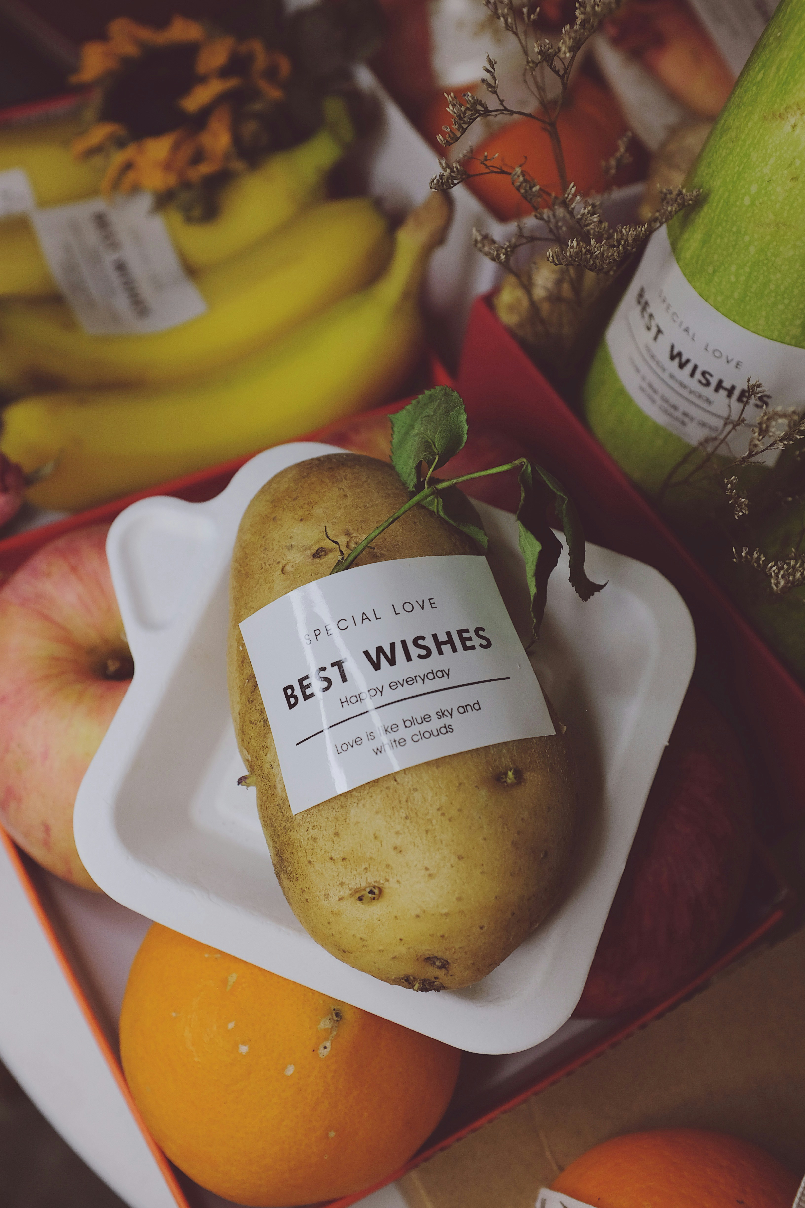 A quirky potato adorned with a label reading 'BEST WISHES' rests on a white plate, surrounded by fresh fruits like apples and oranges. The setting is vibrant and playful.