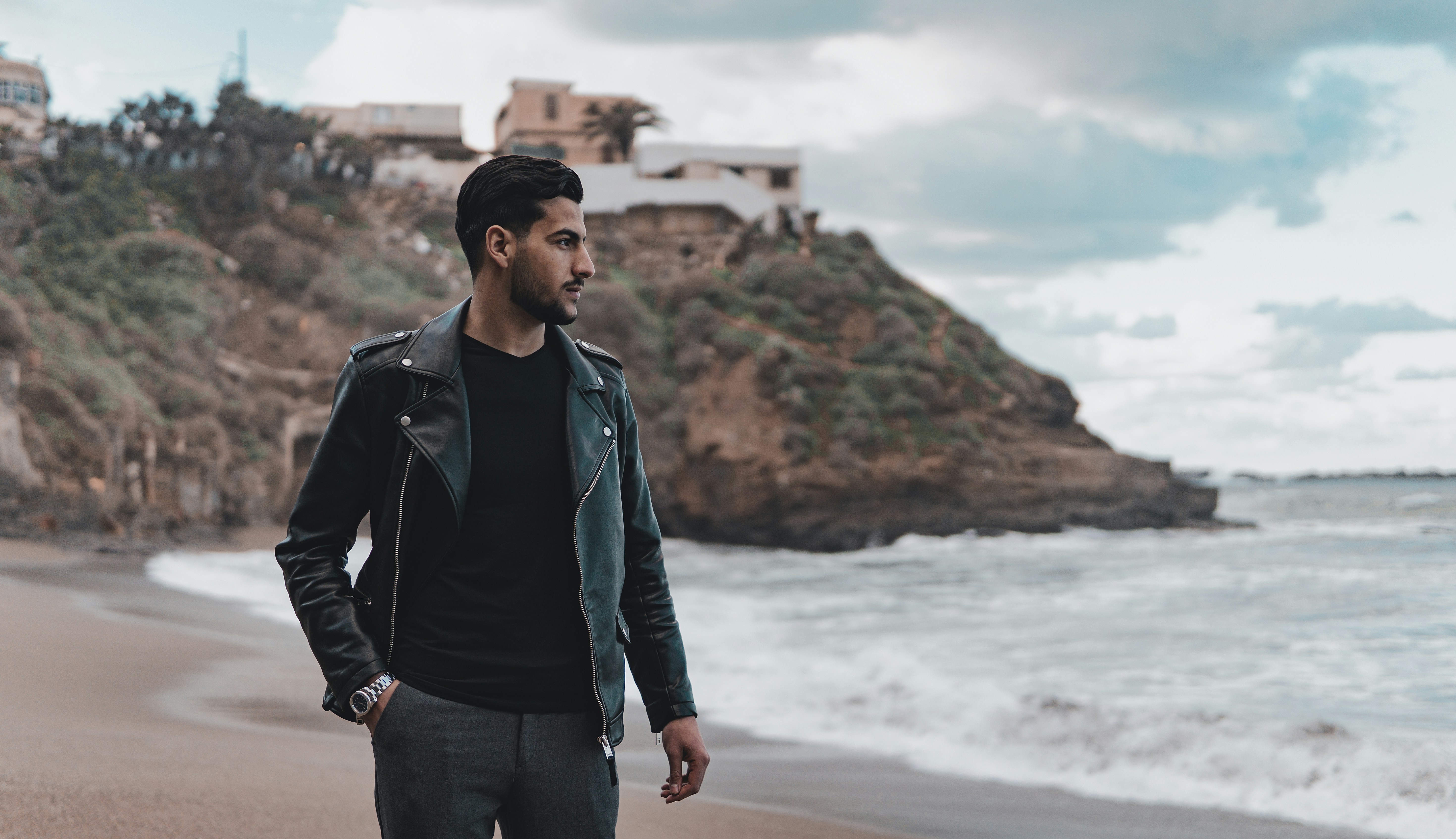 Young man in a leather jacket walking along a sandy beach with rugged cliffs in the background.