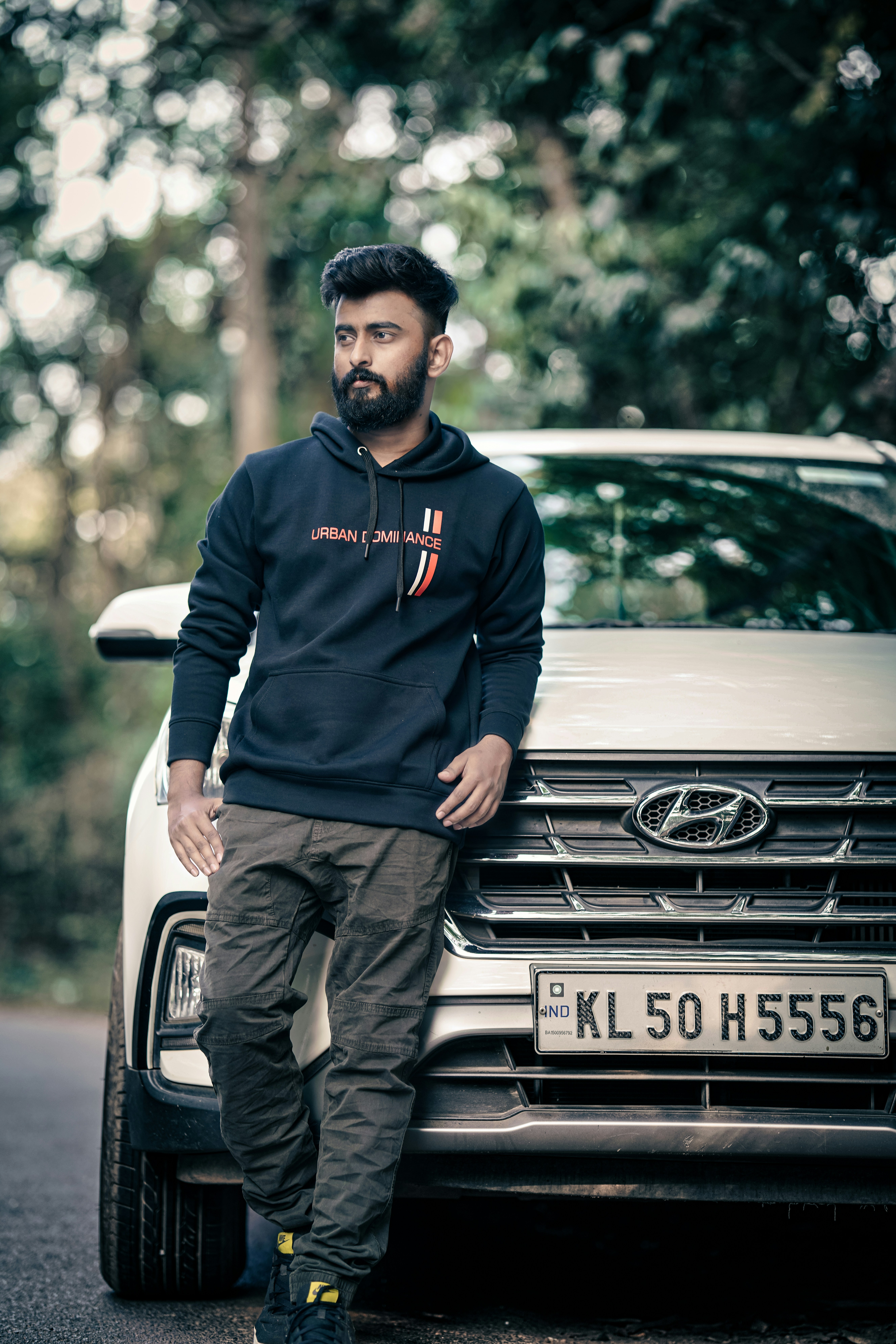 Man in a navy hoodie leaning against a white SUV on a tree-lined road, showcasing a blend of urban style and nature.