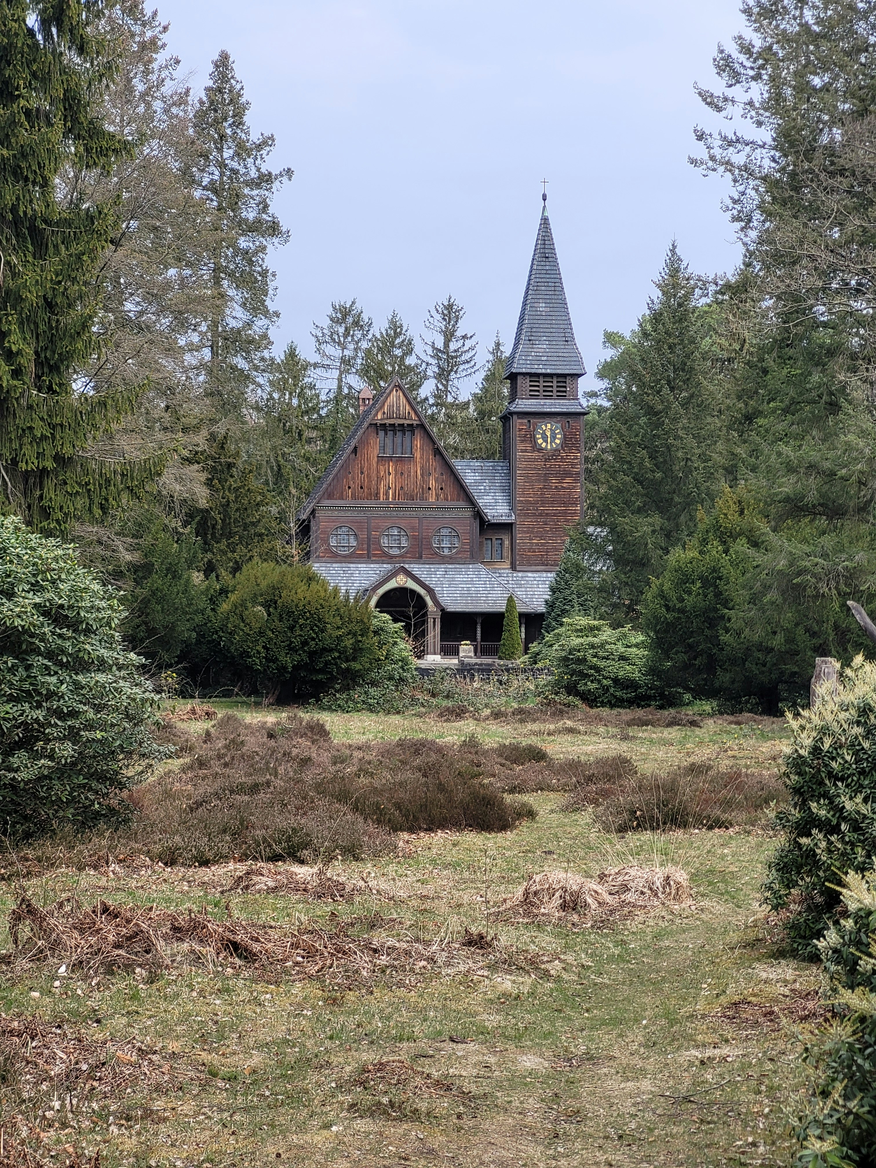 Wooden chapel nestled in a dense forest clearing, surrounded by tall trees and underbrush.