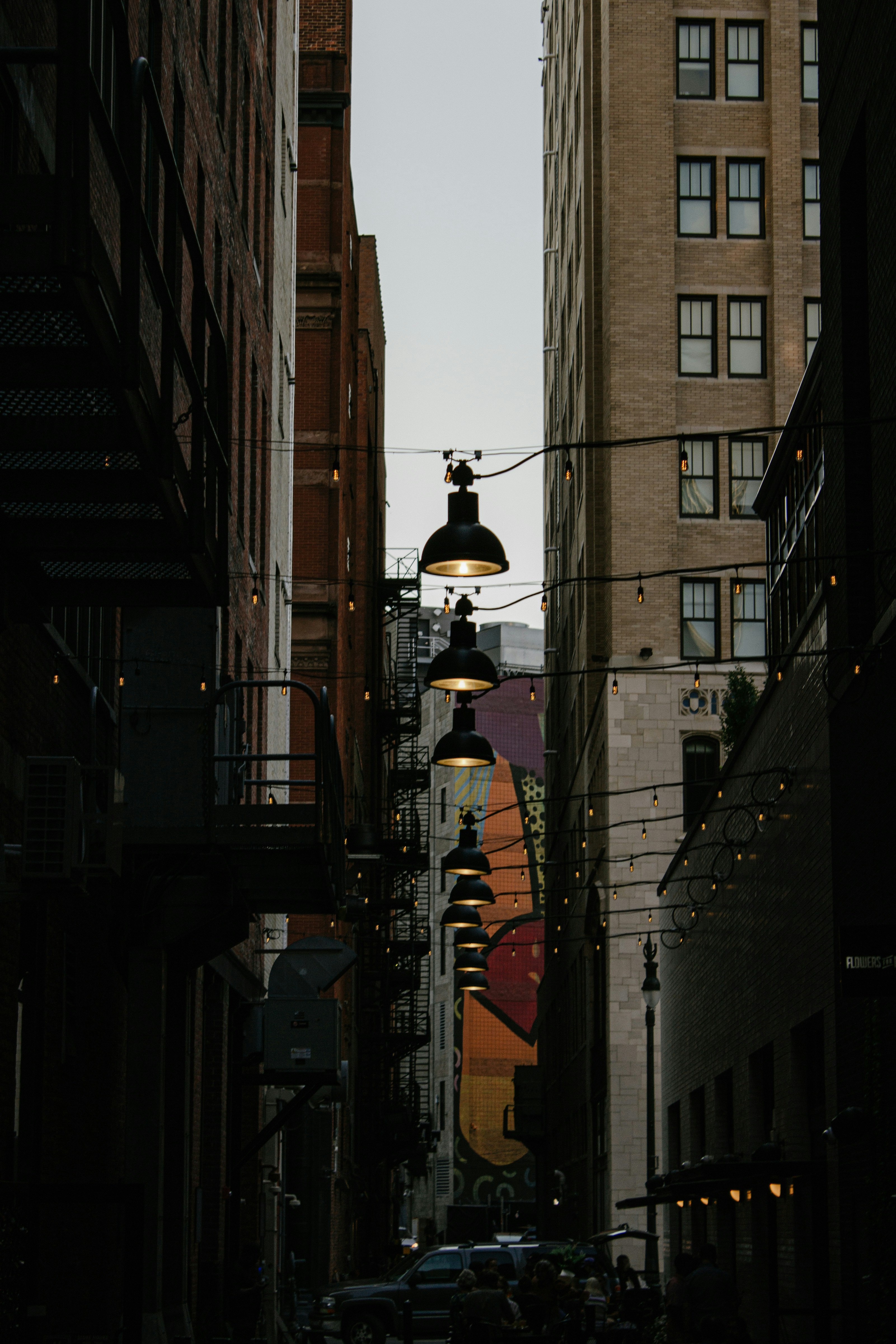 An alleyway adorned with hanging lights, leading to a vibrant mural in the background, showcasing the juxtaposition of urban architecture and artistic expression.