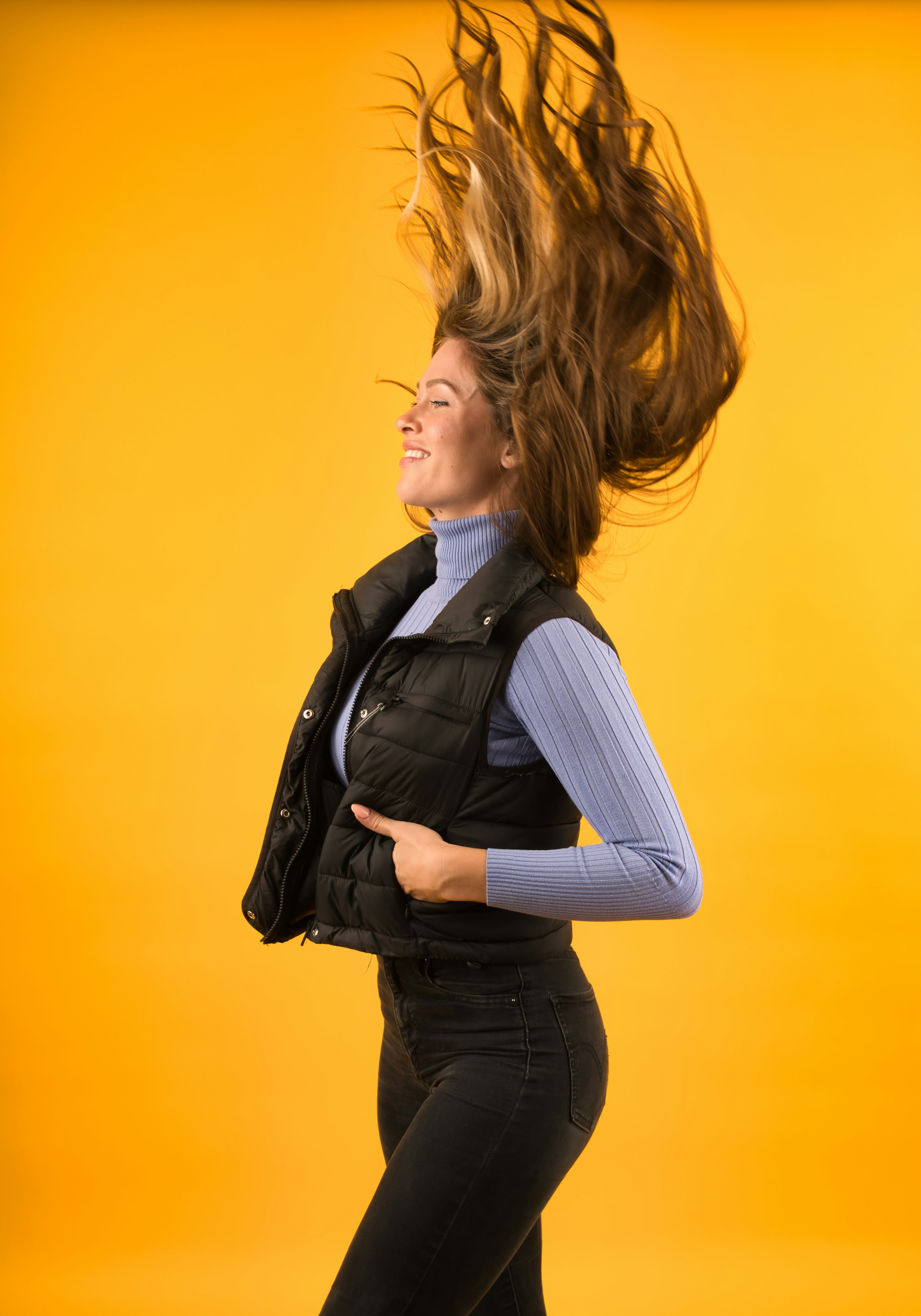 A woman in a black vest and blue turtleneck joyfully flips her hair against a bright yellow background.