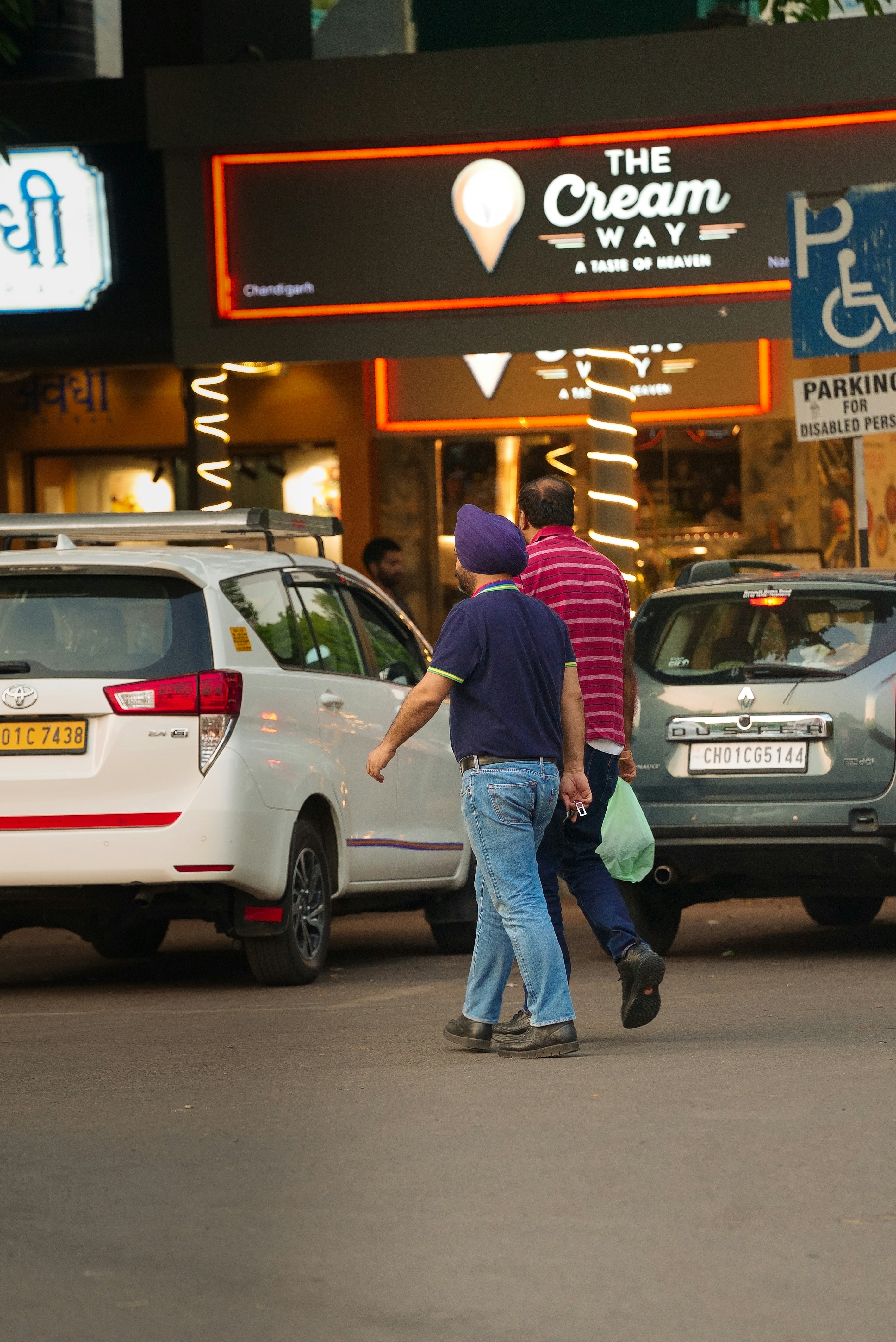 Street photograph of two pedestrians walking away in front of a neon-lit storefront sign reading The Cream Way. Cars line the curb, placing the scene within a busy urban block.
