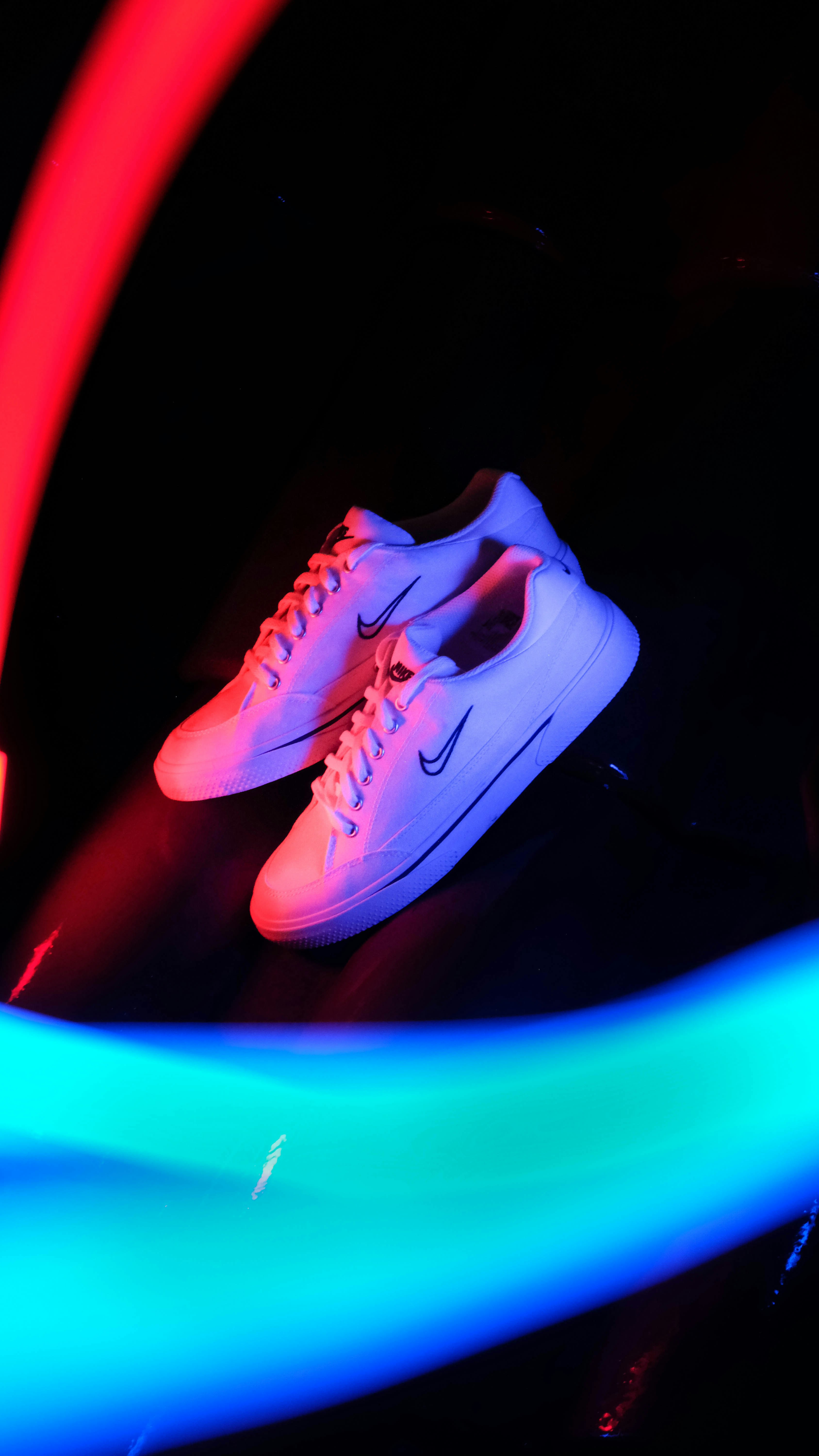 White Nike sneakers illuminated by vibrant red and blue light trails against a dark background.