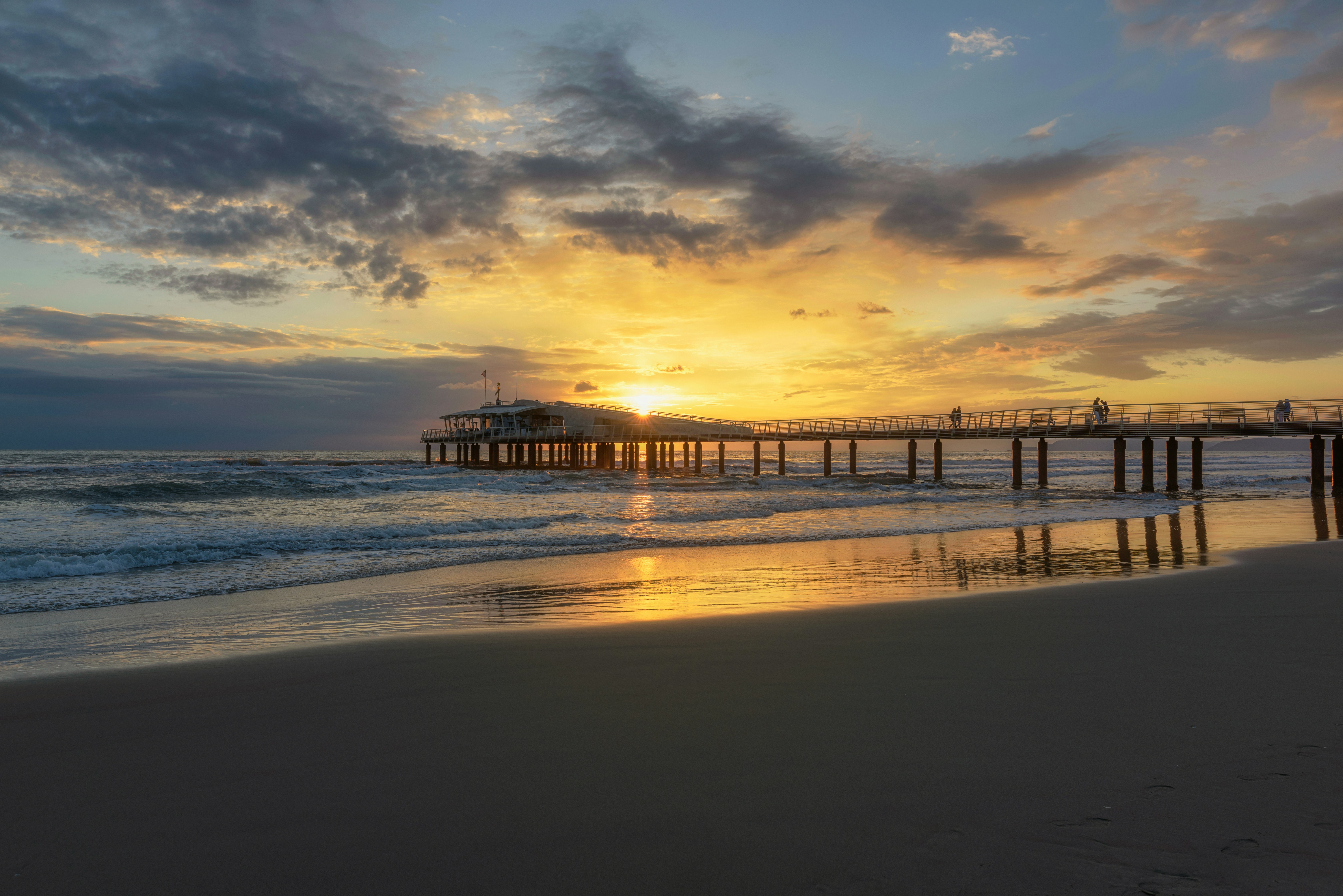 Pier extending into the ocean at sunrise, with vibrant sky reflections on the wet sand.