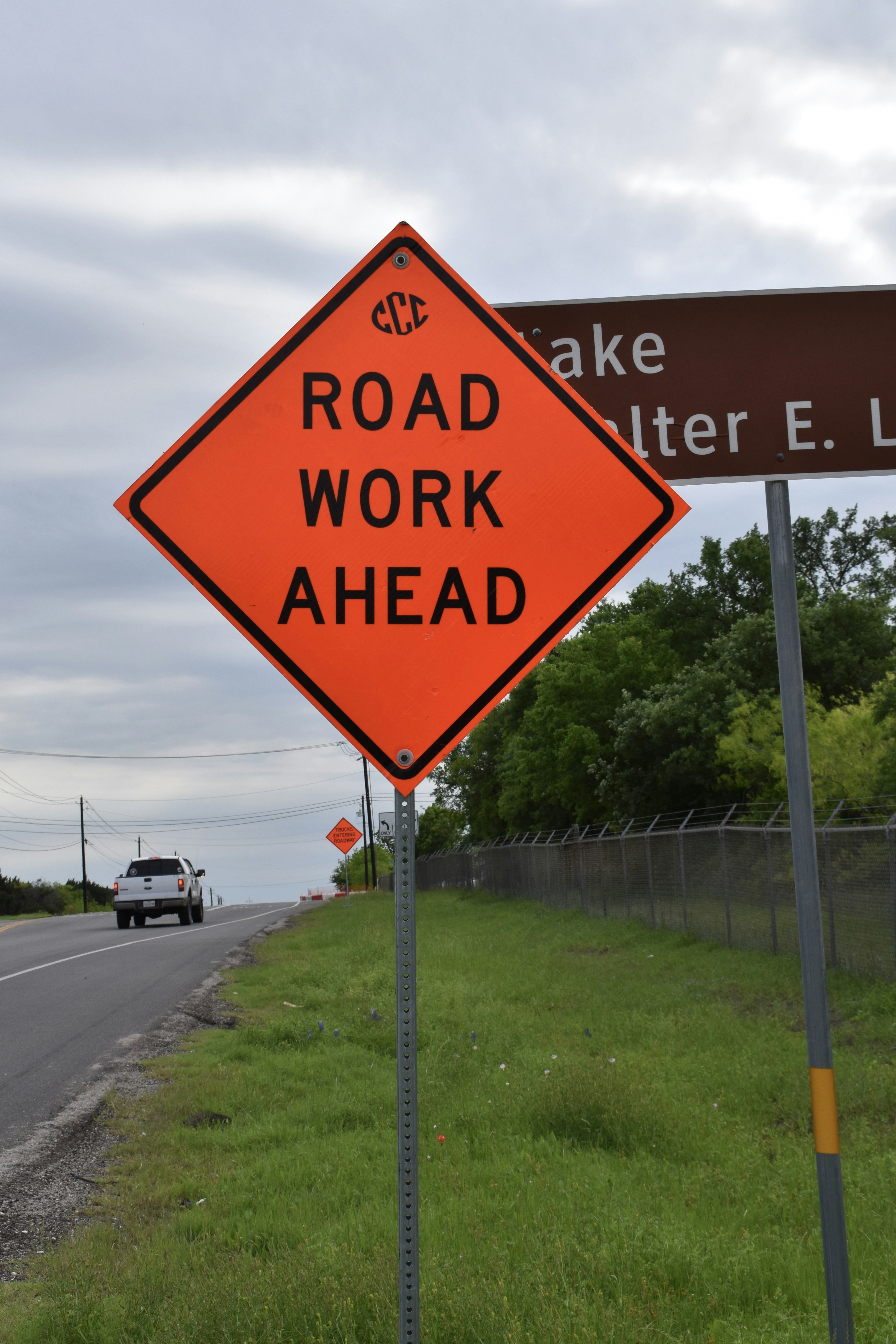 Bright orange road work sign alerts drivers to upcoming construction, set against a cloudy sky and roadside scenery.