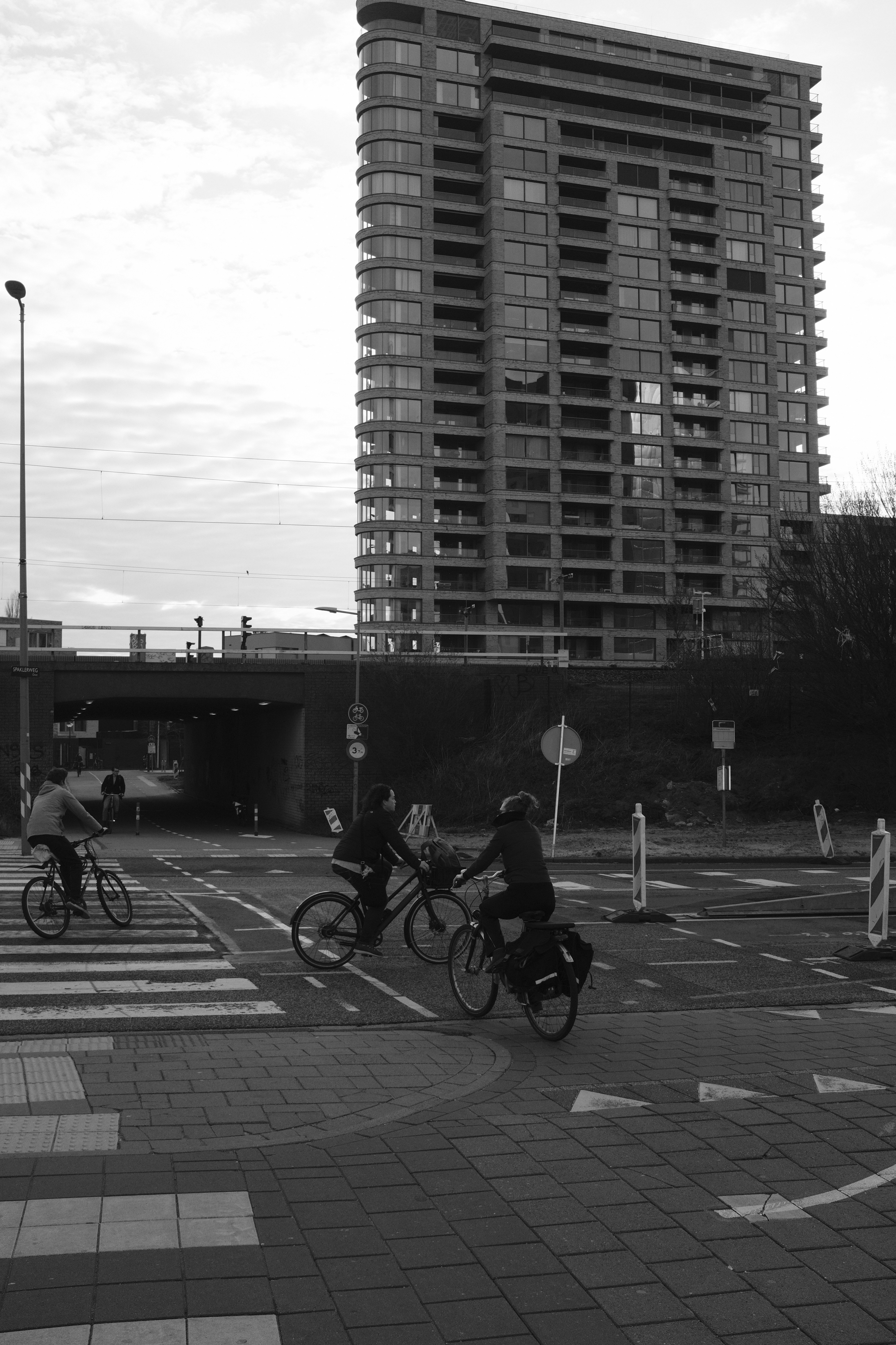 Cyclists navigate a crosswalk in an urban setting, with a contemporary high-rise building in the background. The scene captures the essence of city life and mobility.