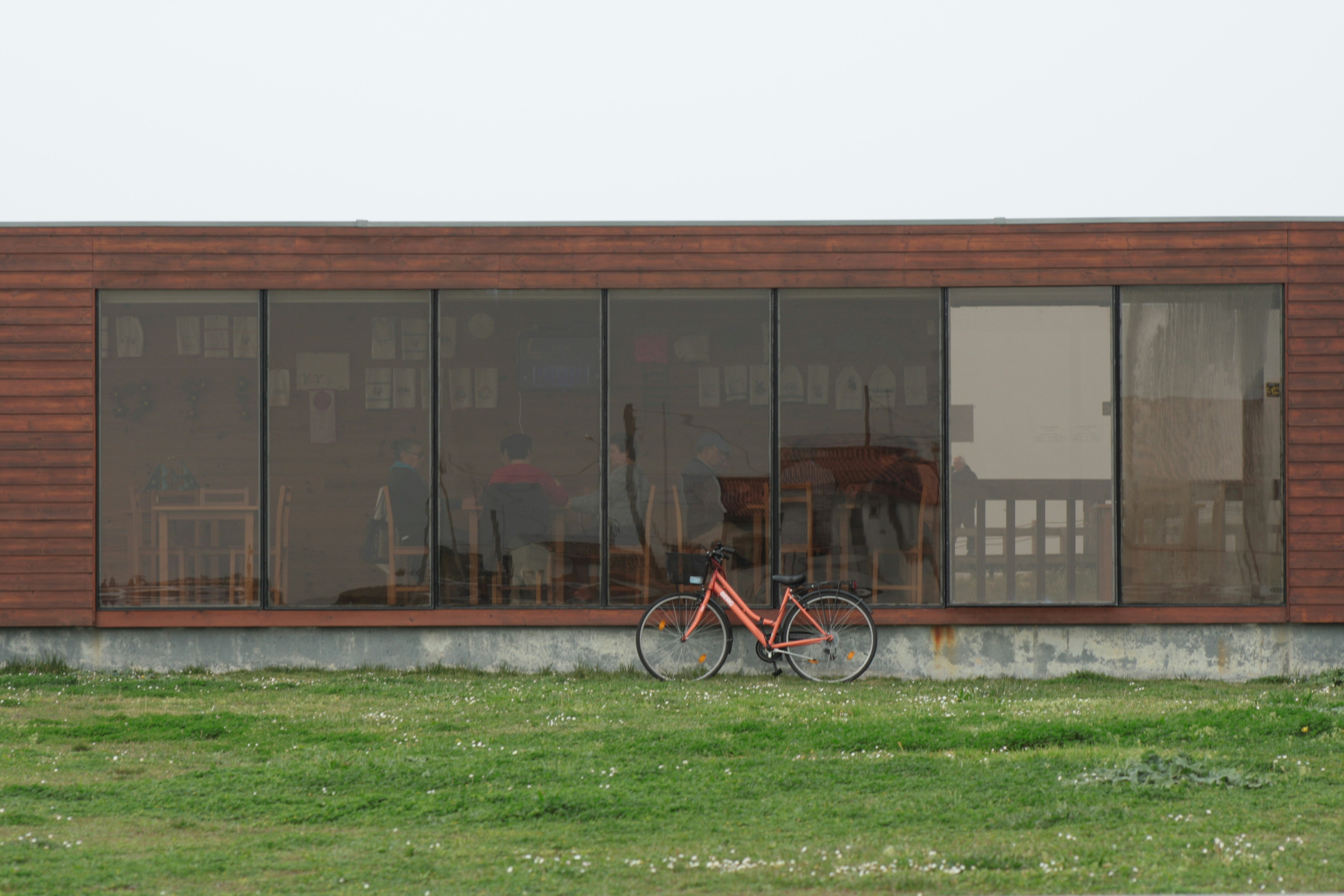 Modern wooden building with large glass windows, revealing a cozy interior, accompanied by a parked orange bicycle on a grassy field.