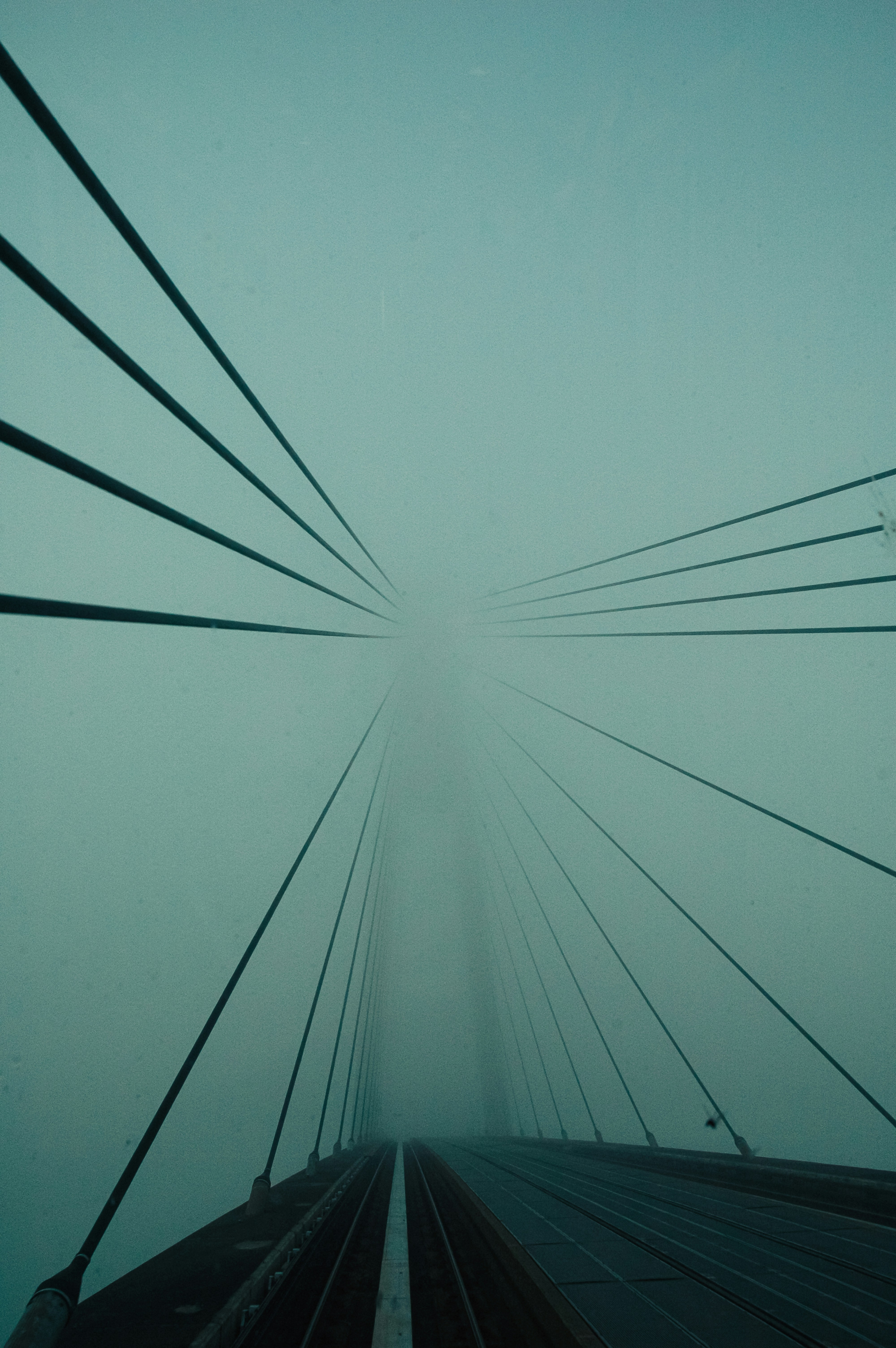 Suspension bridge disappearing into thick fog, creating an eerie atmosphere. The cables stretch toward the obscured horizon.