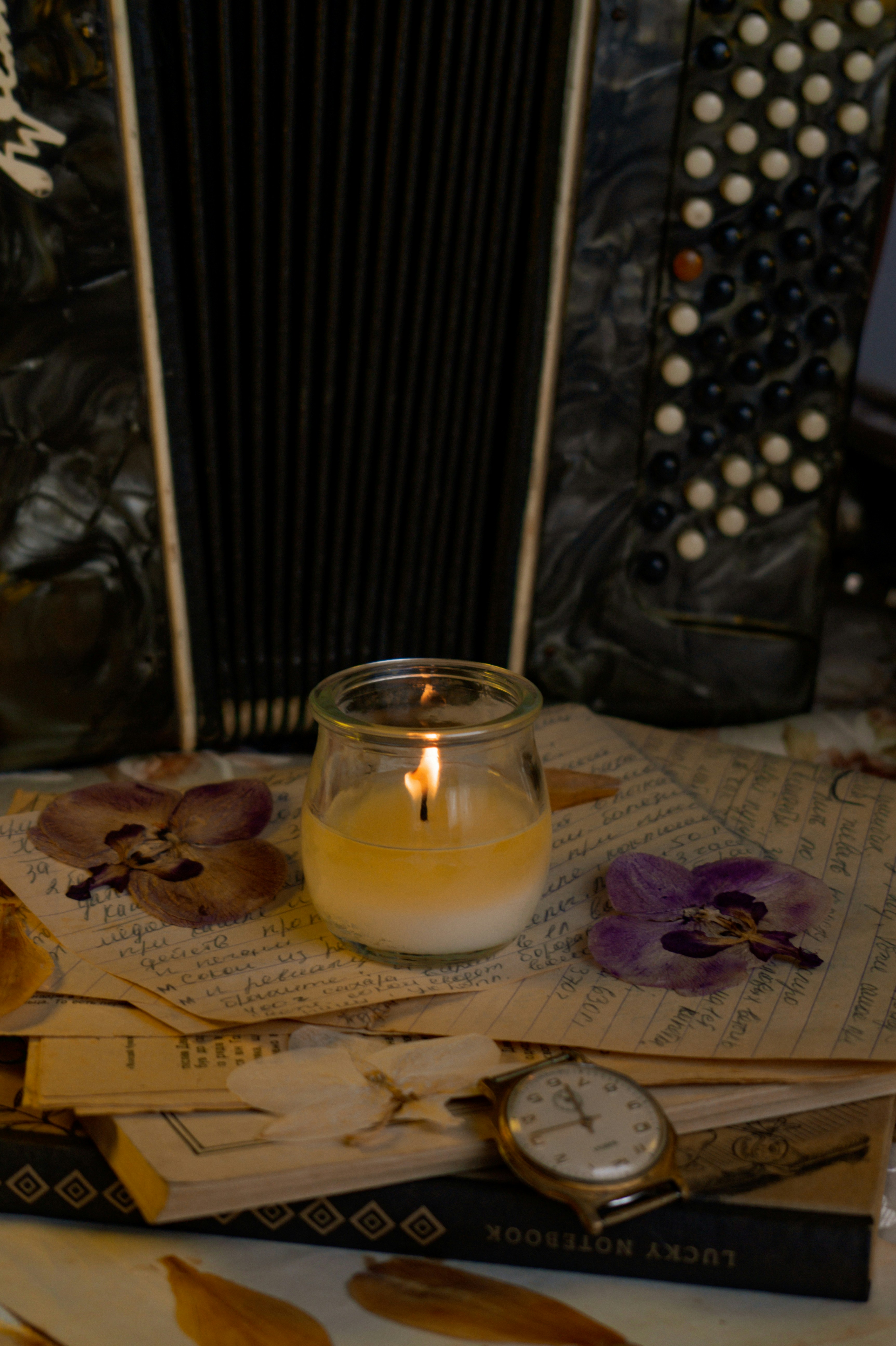 A warm candle glows in a glass jar atop handwritten papers and a vintage pocket watch. An aged accordion backdrop adds depth to this moody still-life photograph.