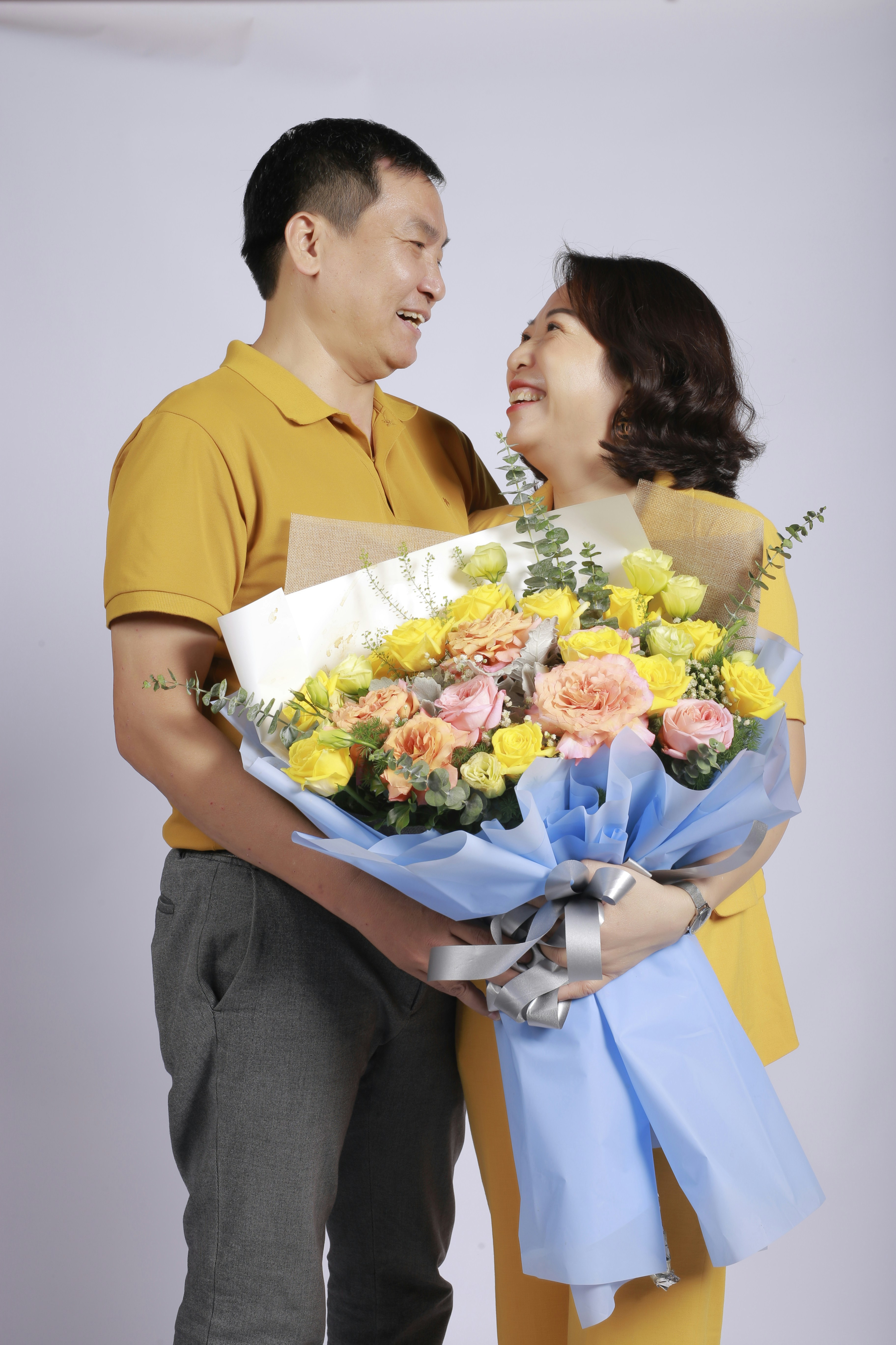 Couple sharing a joyful moment while holding a large bouquet of colorful flowers against a soft backdrop.