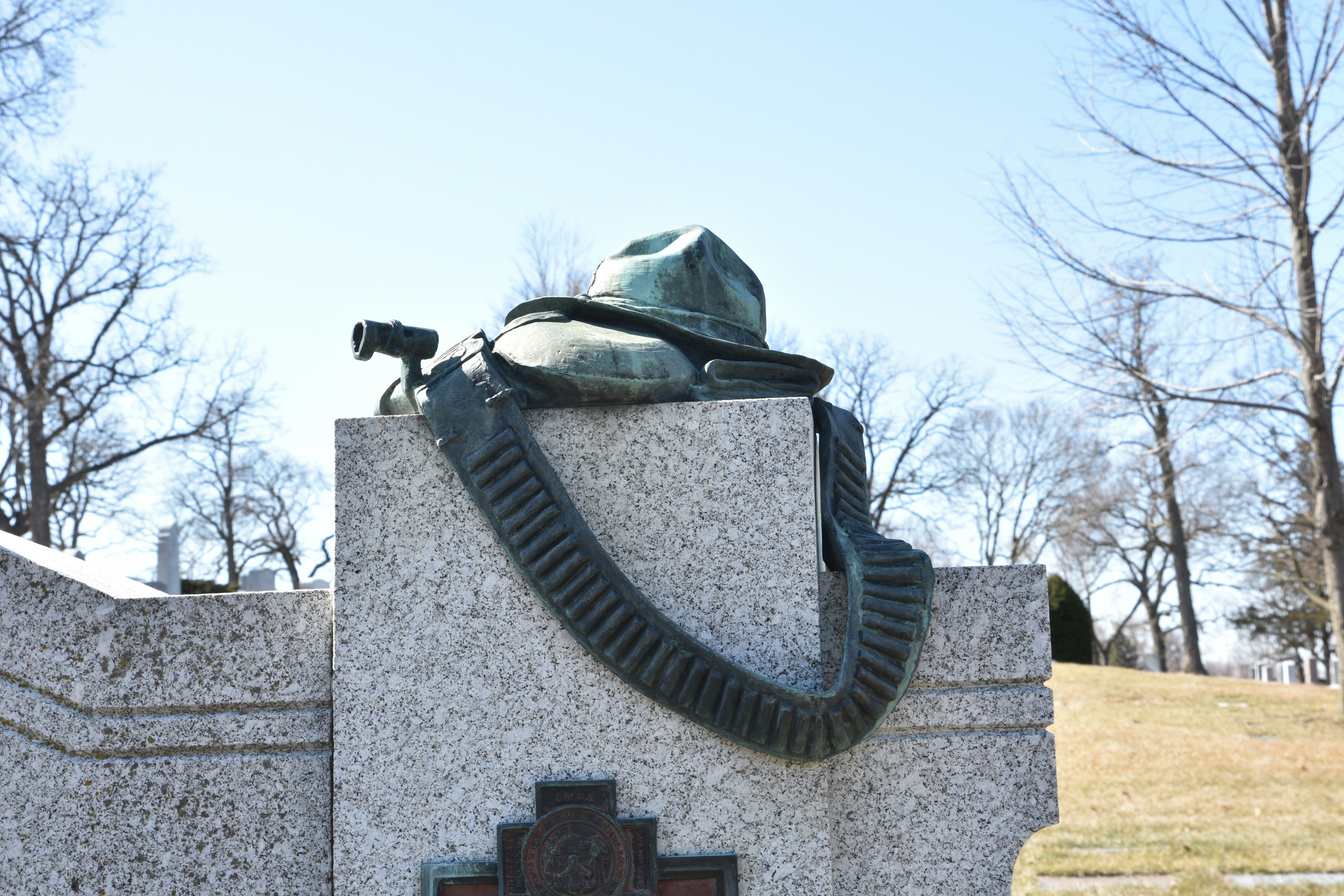 Bronze military helmet resting on a gravestone, accompanied by a decorative belt, set against a serene backdrop of trees.