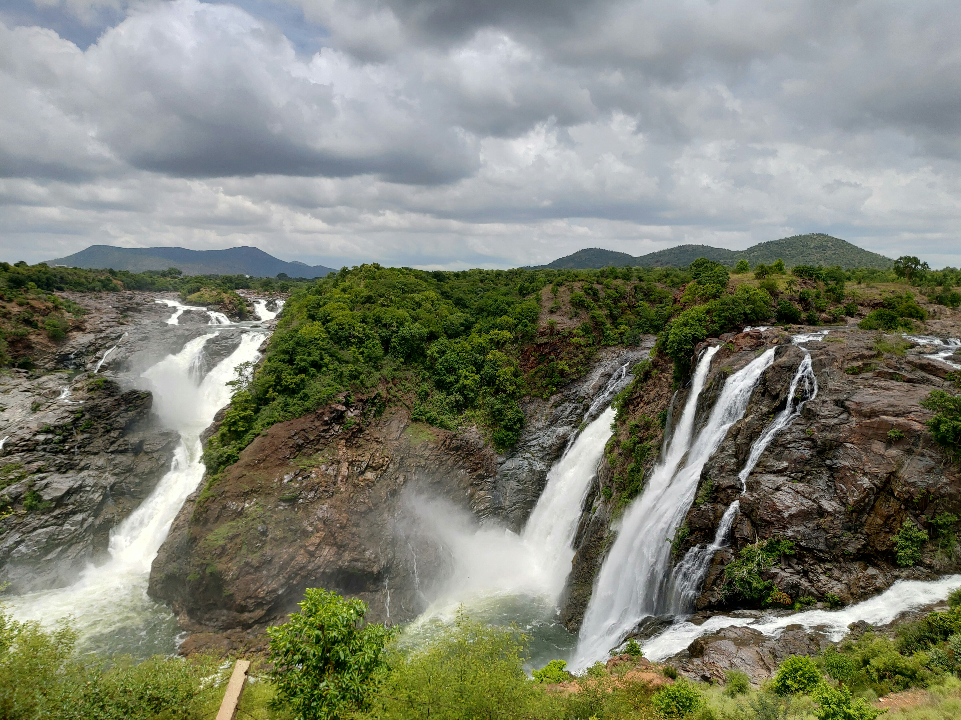 Majestic waterfalls cascading down rocky cliffs, surrounded by lush greenery and distant hills under a cloudy sky.