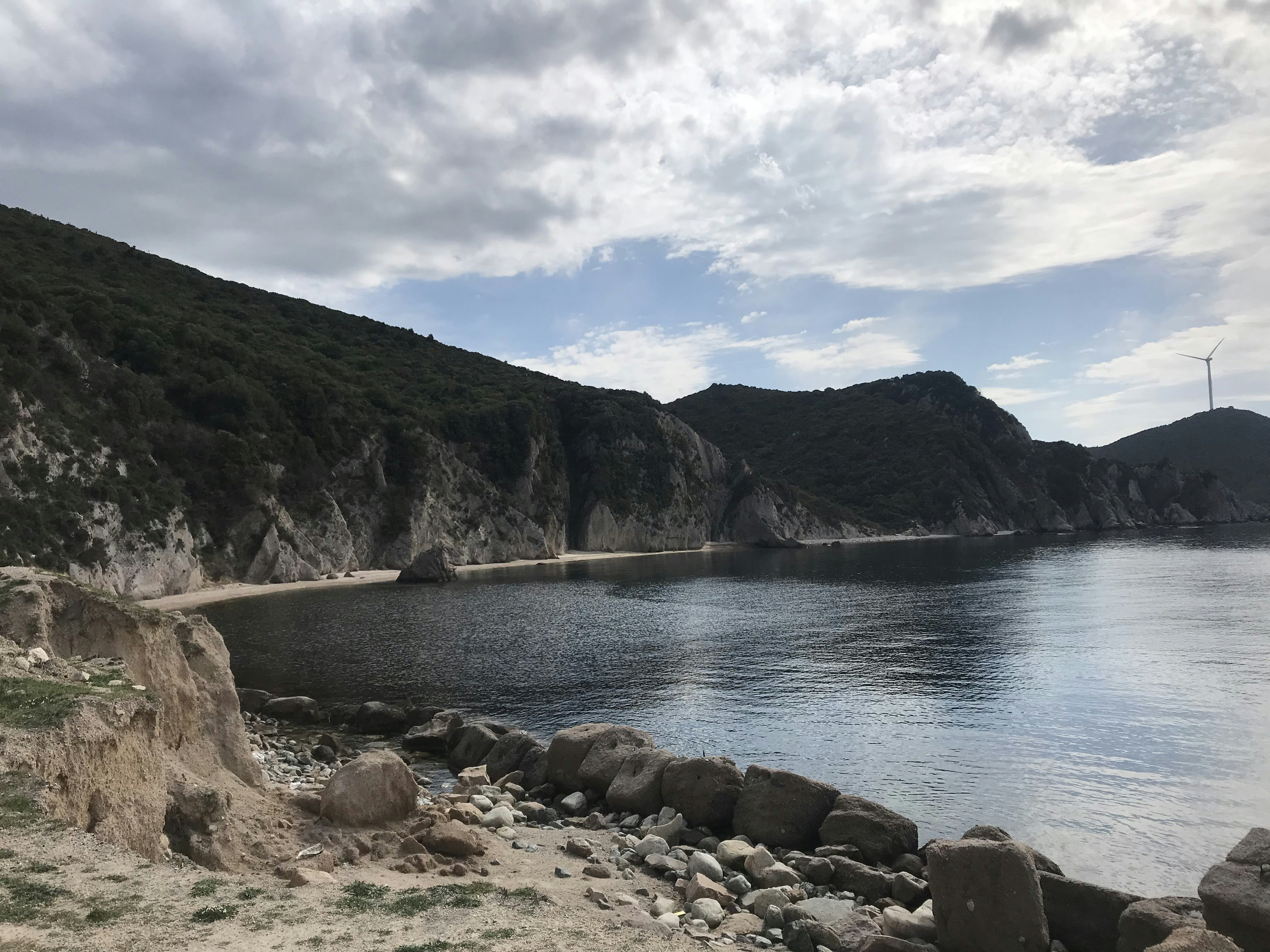 Calm body of water bordered by rocky cliffs and lush mountains under a cloudy sky.