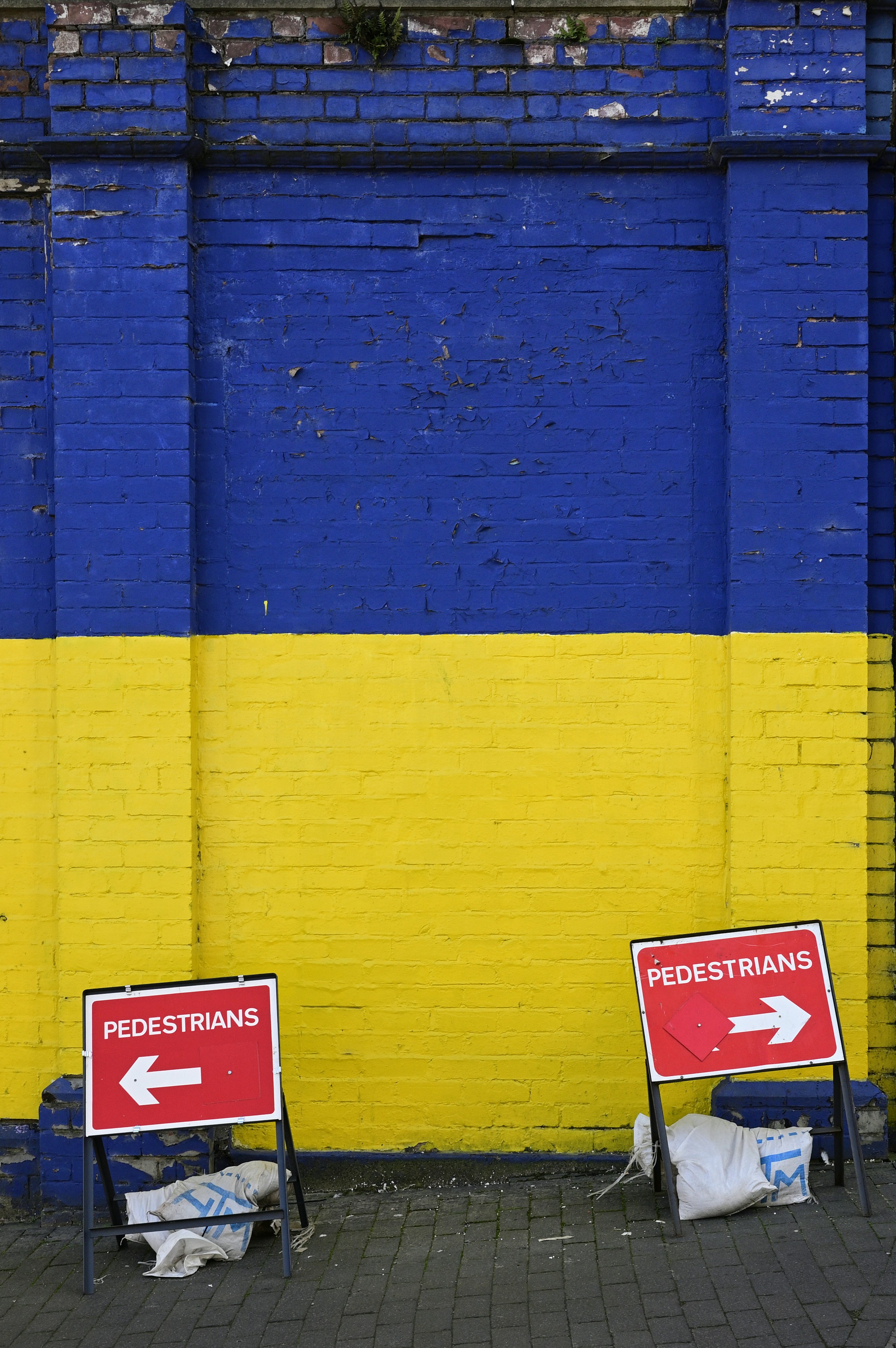 Blue-Gold Street Palette: Pedestrian Signs Framing a Color-Blocked Brick Wall