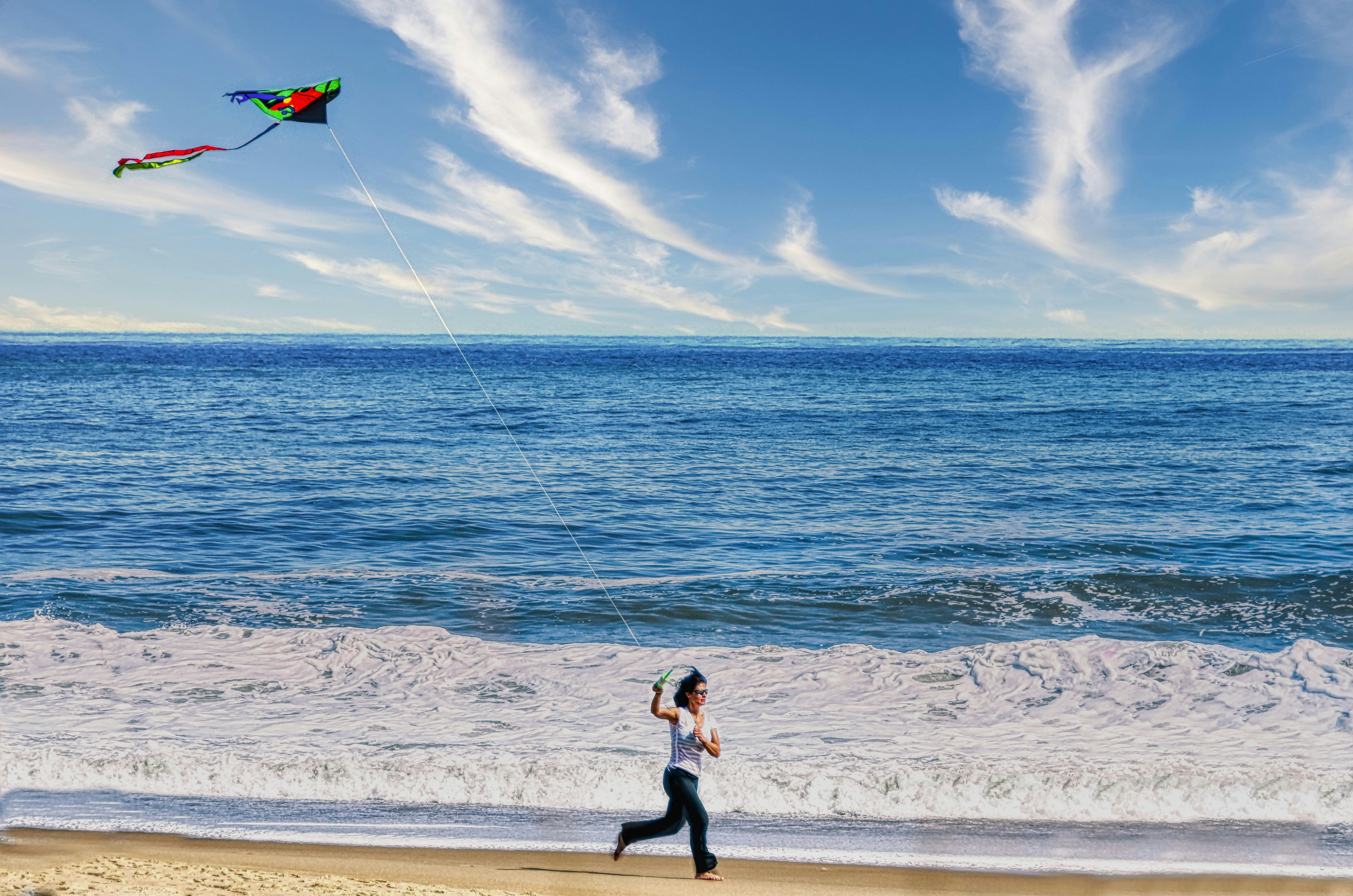A person joyfully runs along the beach, flying a vibrant kite against a backdrop of serene ocean waves and a bright sky.