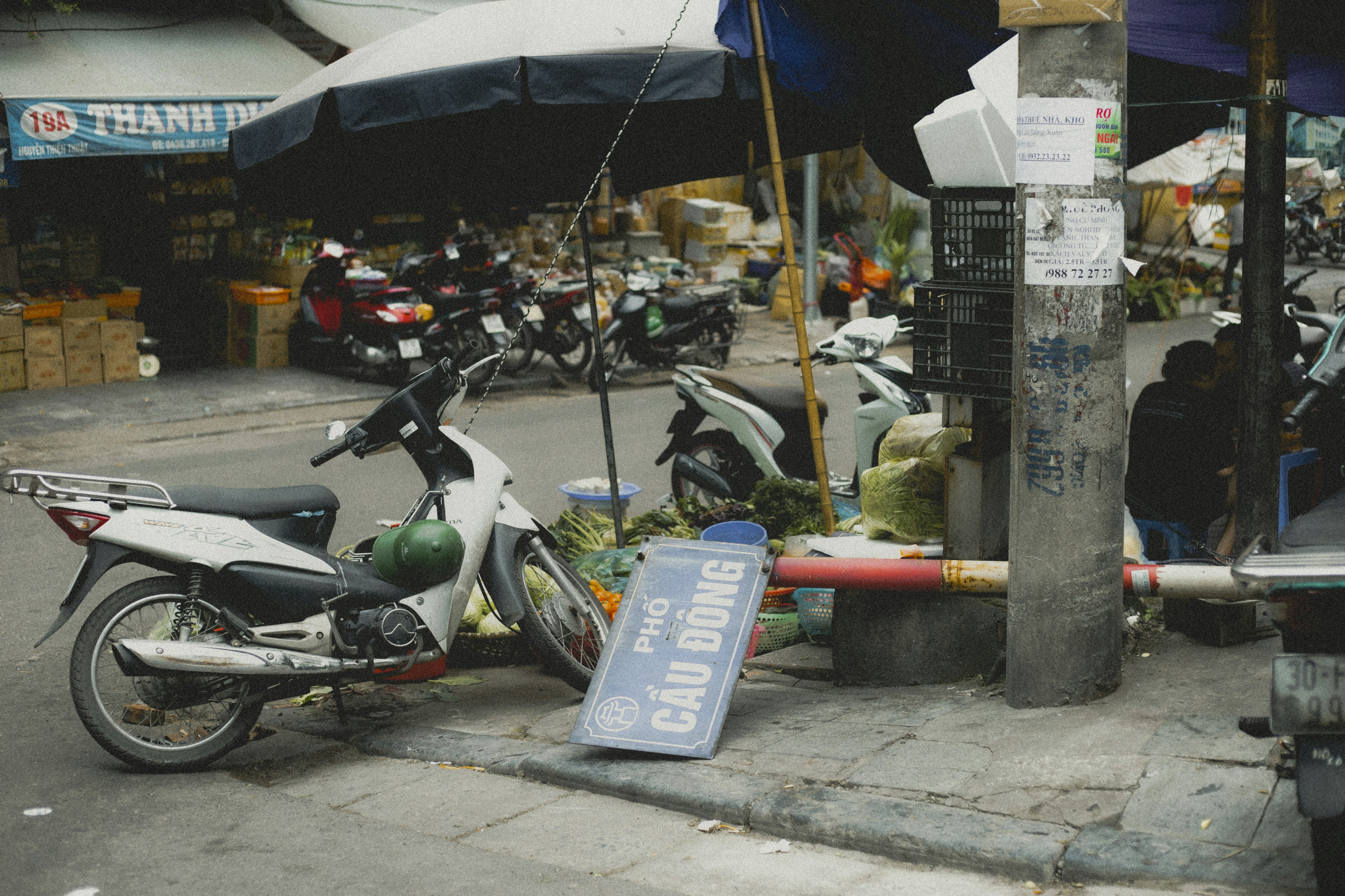A motorbike parked beside a street vendor's stall, with a sign reading 'Phở Cầu Đồng' leaning against a pole in a bustling market area.
