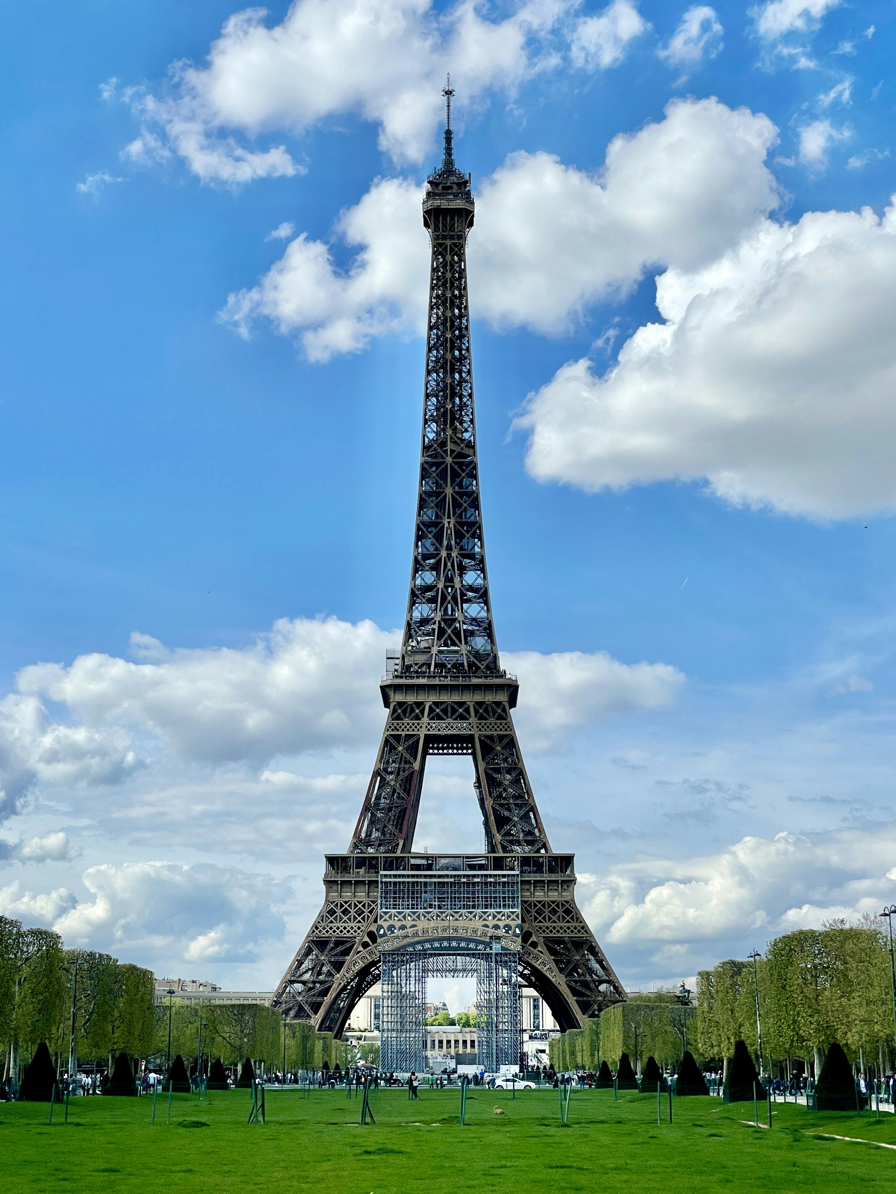 Eiffel Tower rising majestically against a backdrop of fluffy clouds in a vibrant blue sky, framed by manicured green lawns and trees.