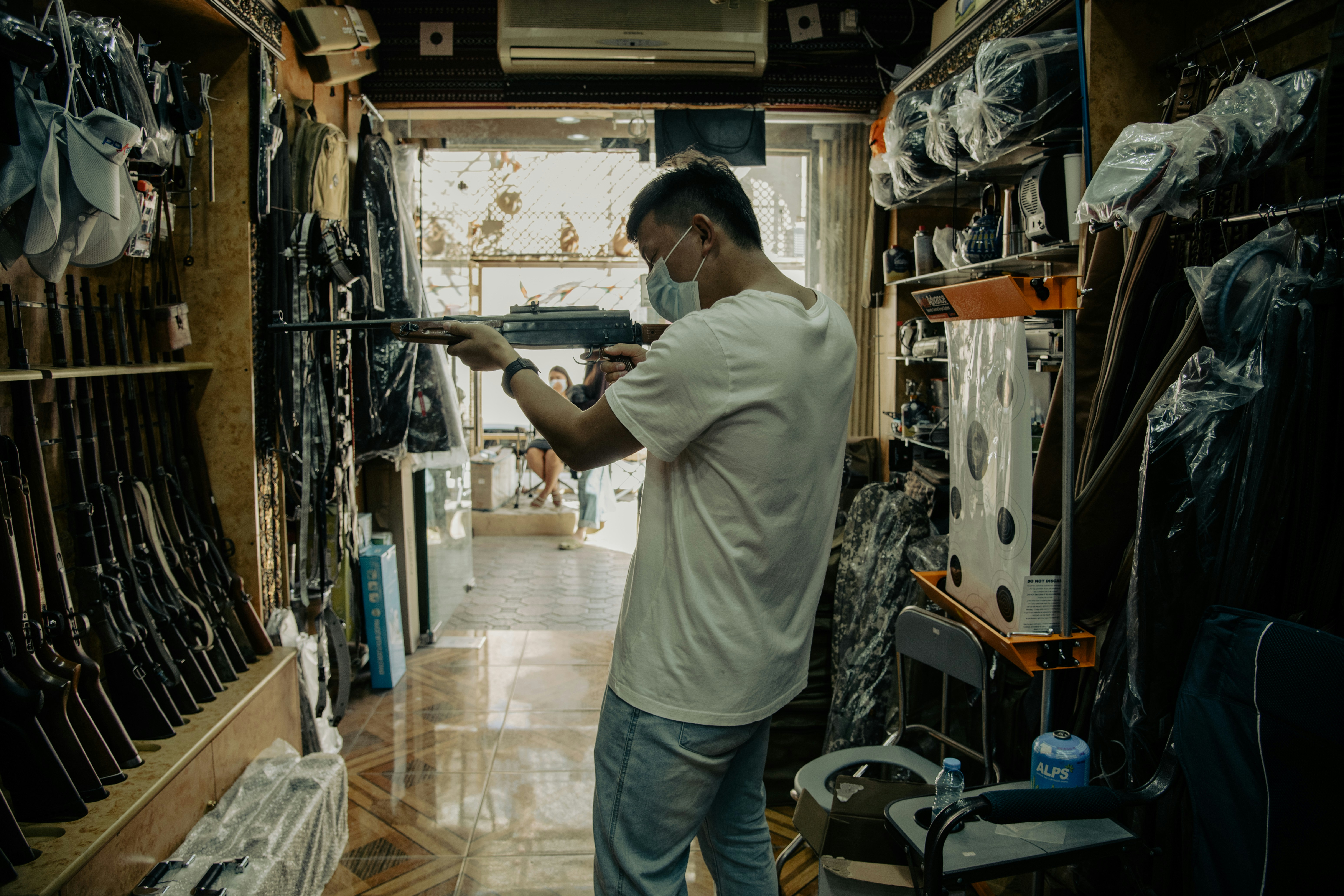 A person aiming a firearm in a shop filled with various weapons and accessories. The setting reflects a specialized environment for enthusiasts.