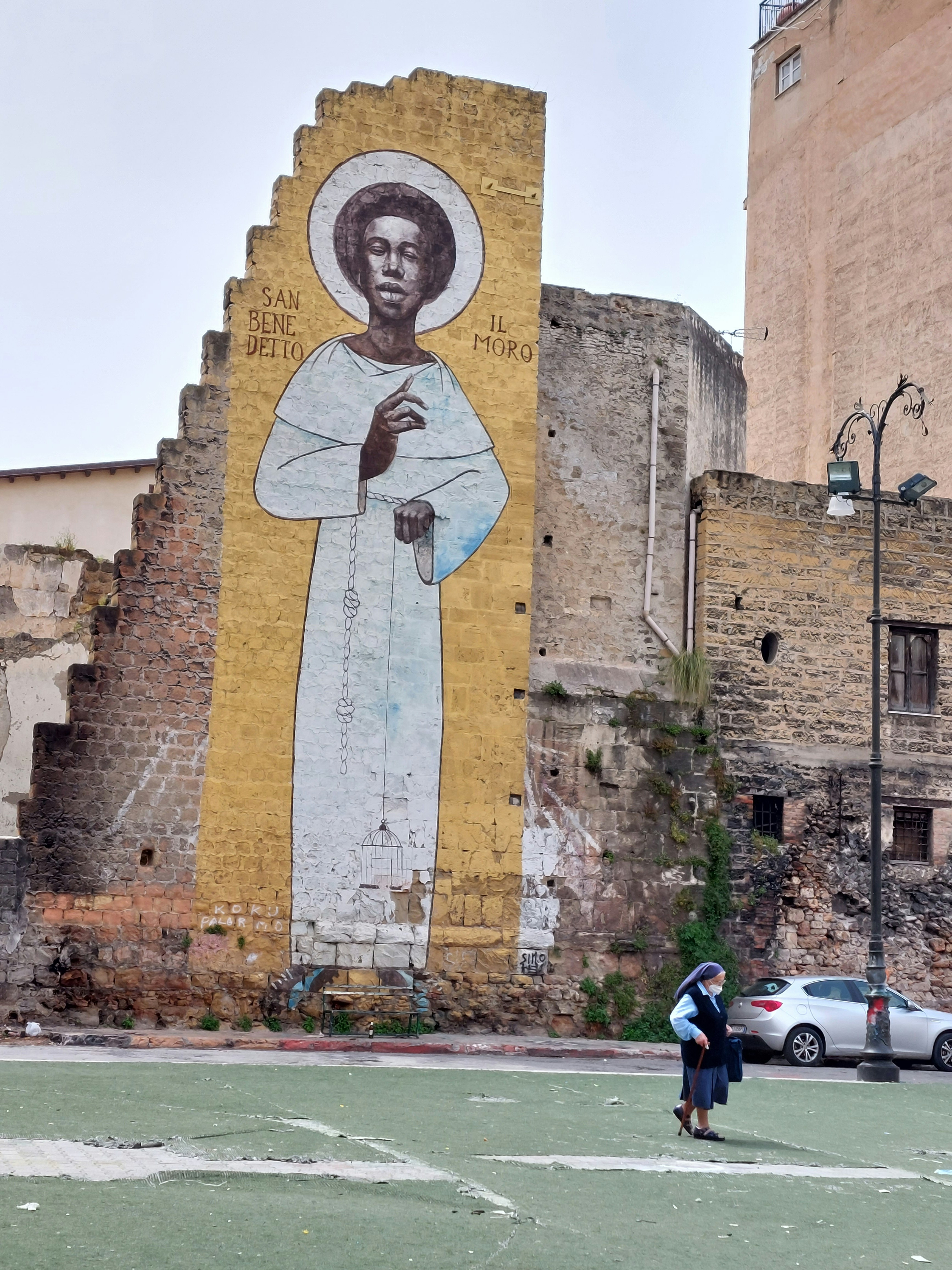 Large mural painting of a saint on a weathered brick wall dominates a quiet street, with a woman walking in the foreground for scale.
