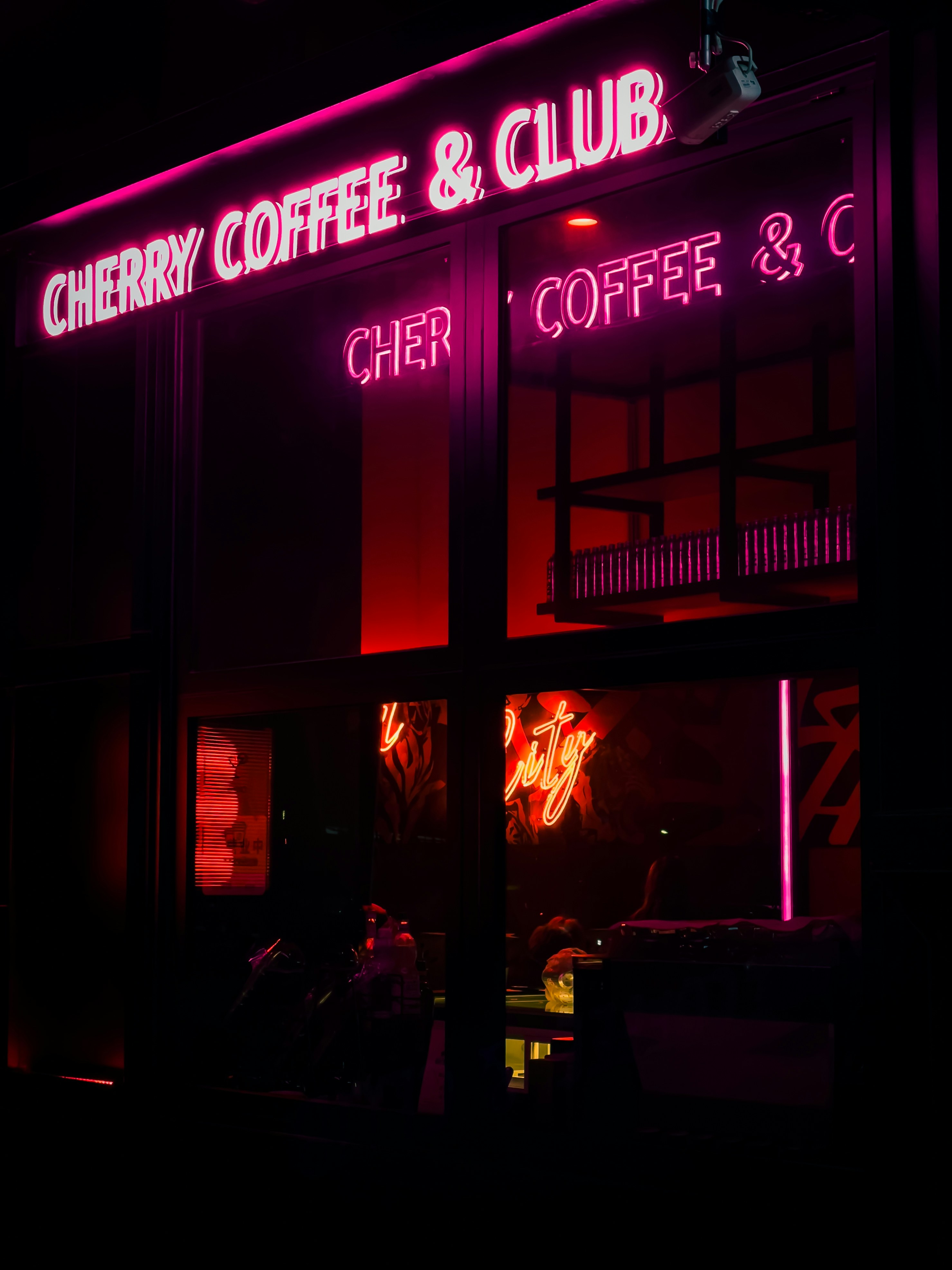 Illuminated storefront of Cherry Coffee & Club featuring vibrant neon signage against a dark backdrop. The interior hints at a lively ambiance.