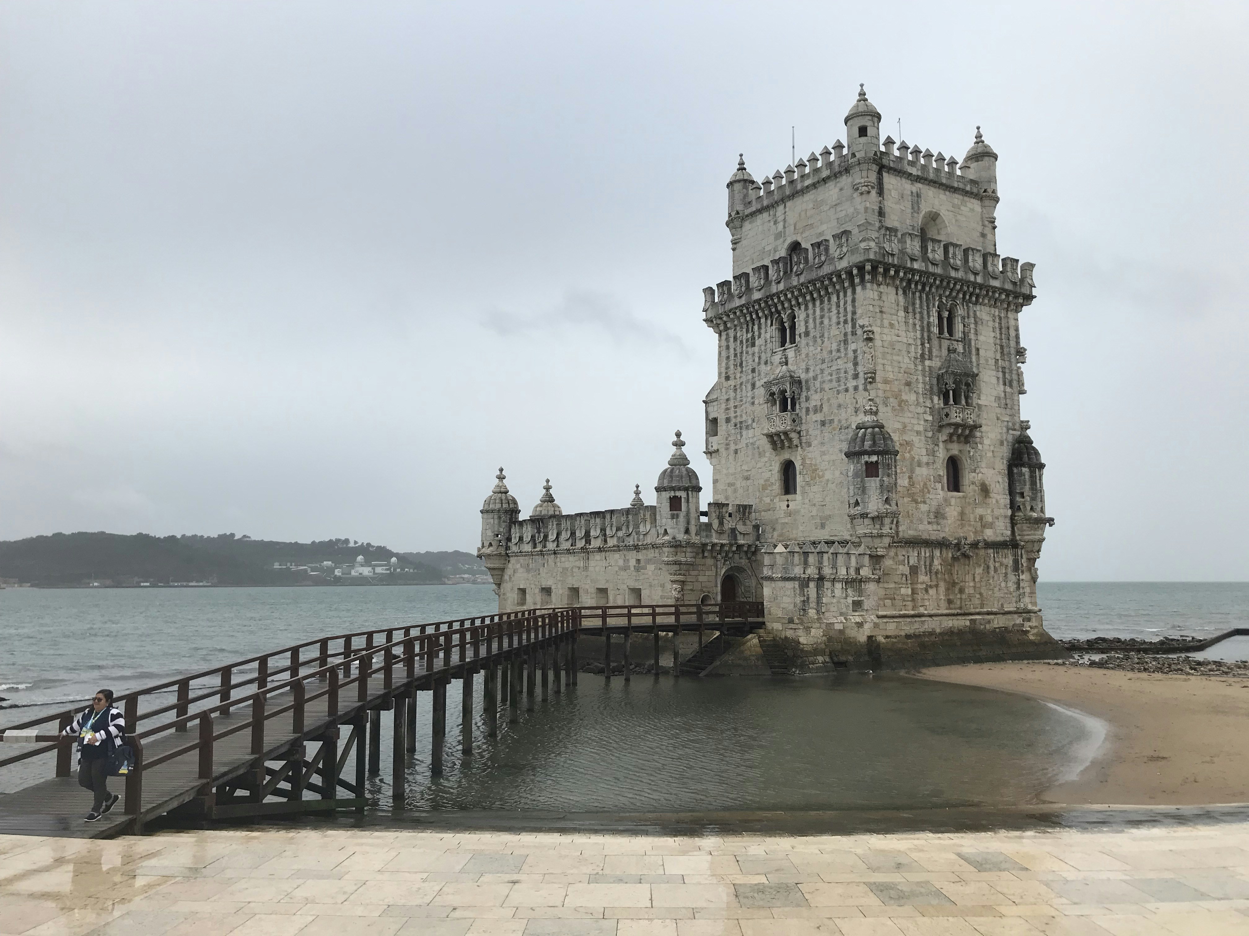Stone tower with battlements stands beside a calm body of water under an overcast sky.