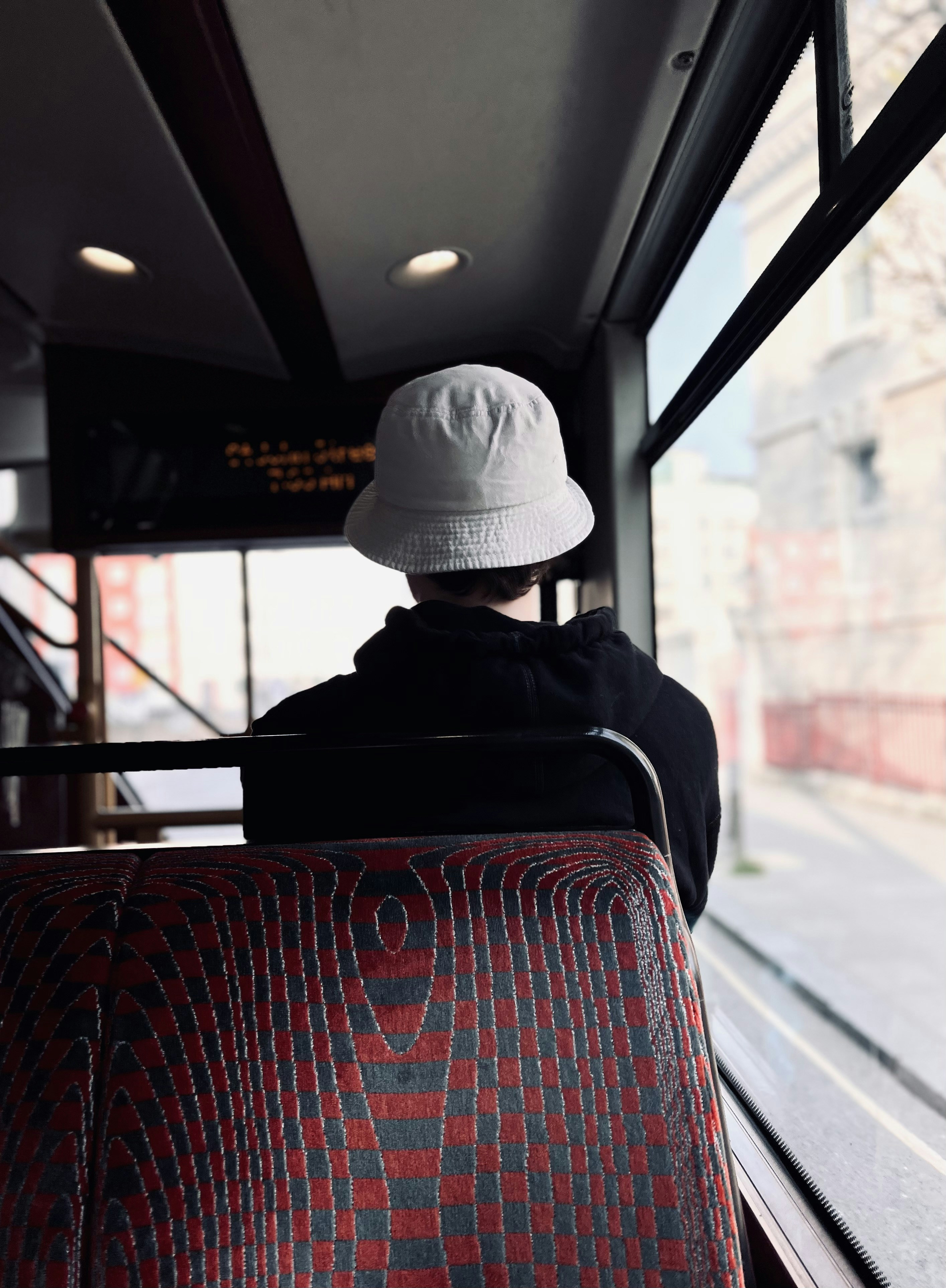 A person wearing a white bucket hat sits on a bus, gazing out the window at the passing urban landscape.