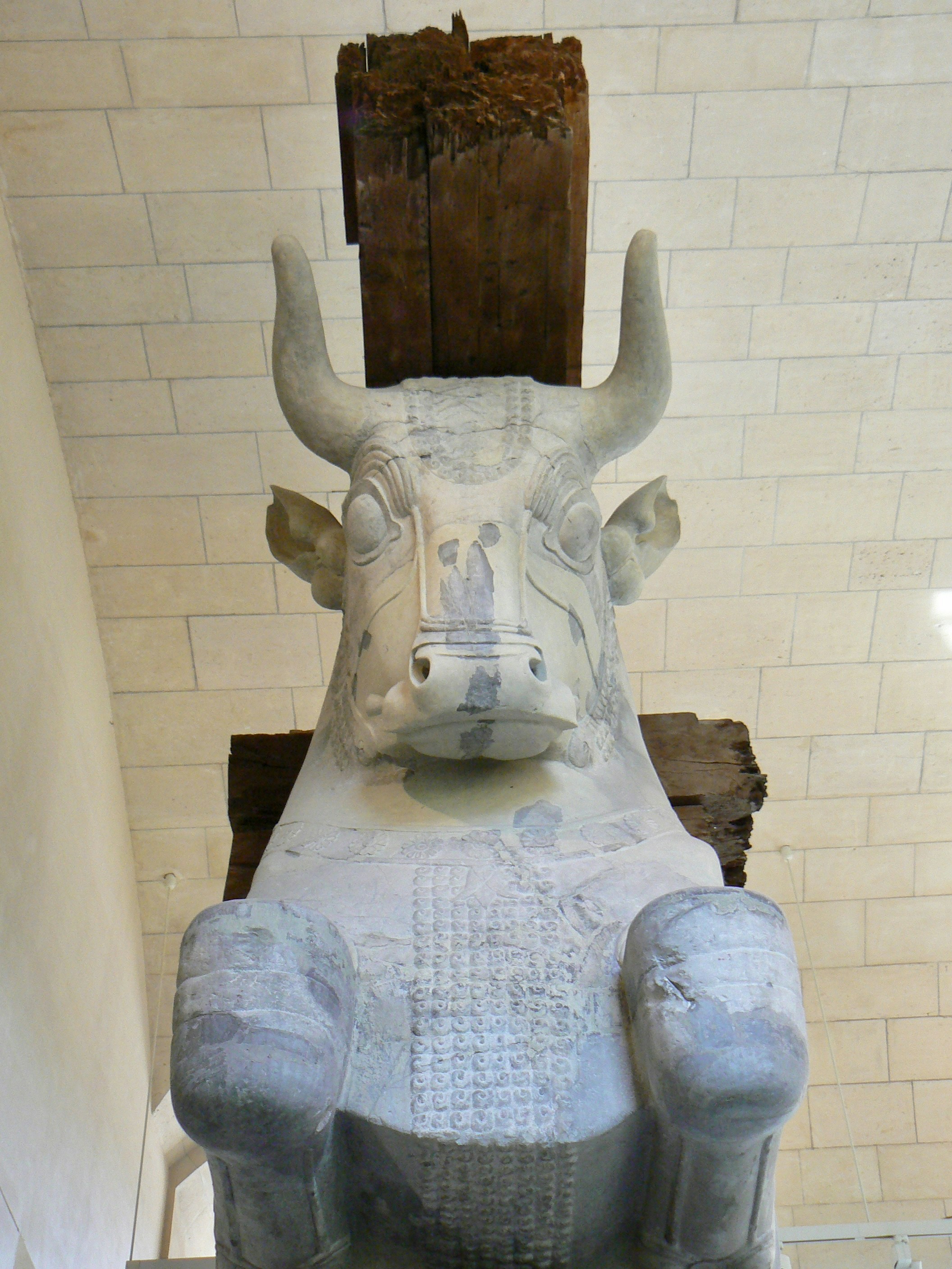 Front view of a large stone bull statue with raised forelegs in a light-tiled hall; a carved guardian beneath a wooden beam.