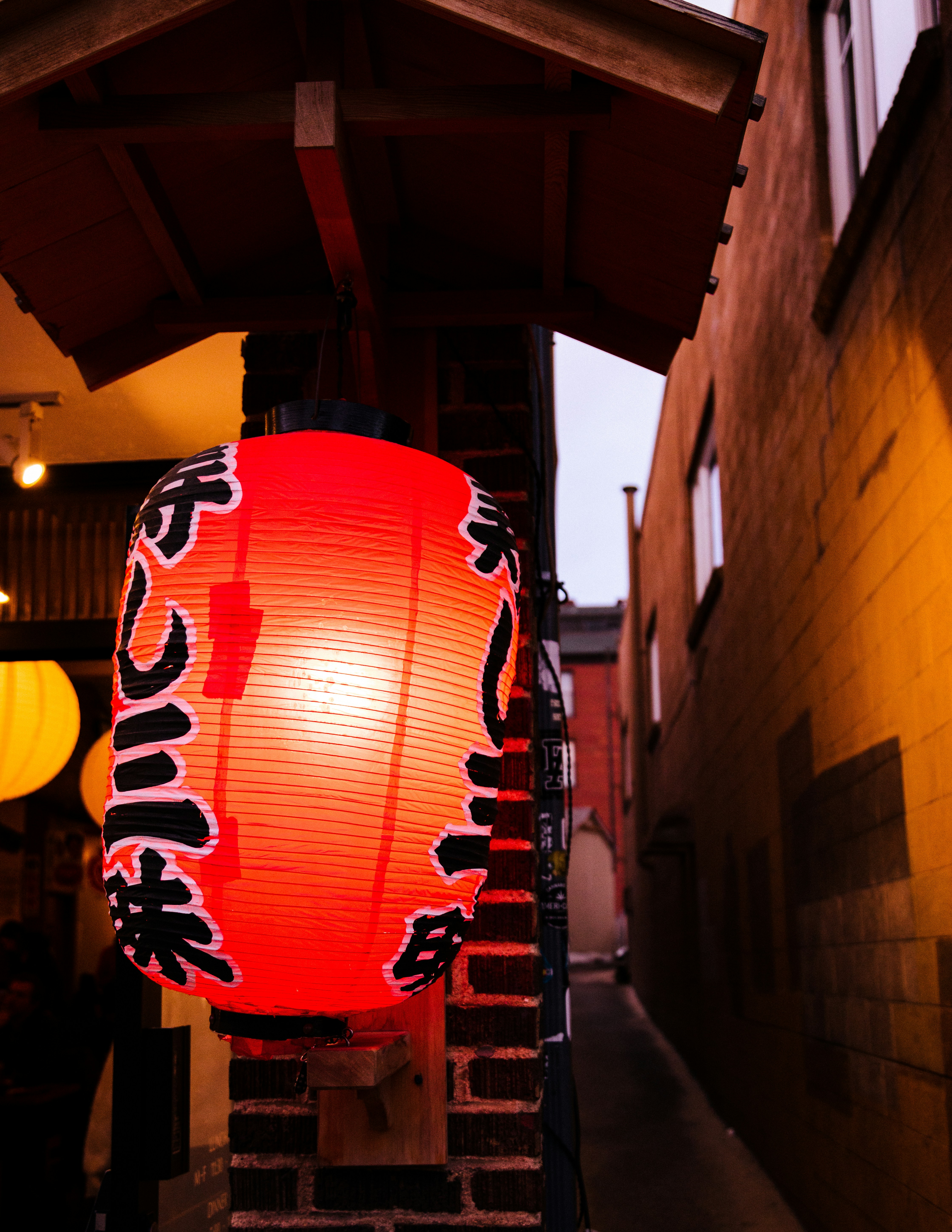 Vibrant red lantern hanging in a narrow alley, casting warm light against the brick wall. The intricate characters adorn the lantern, adding cultural depth.