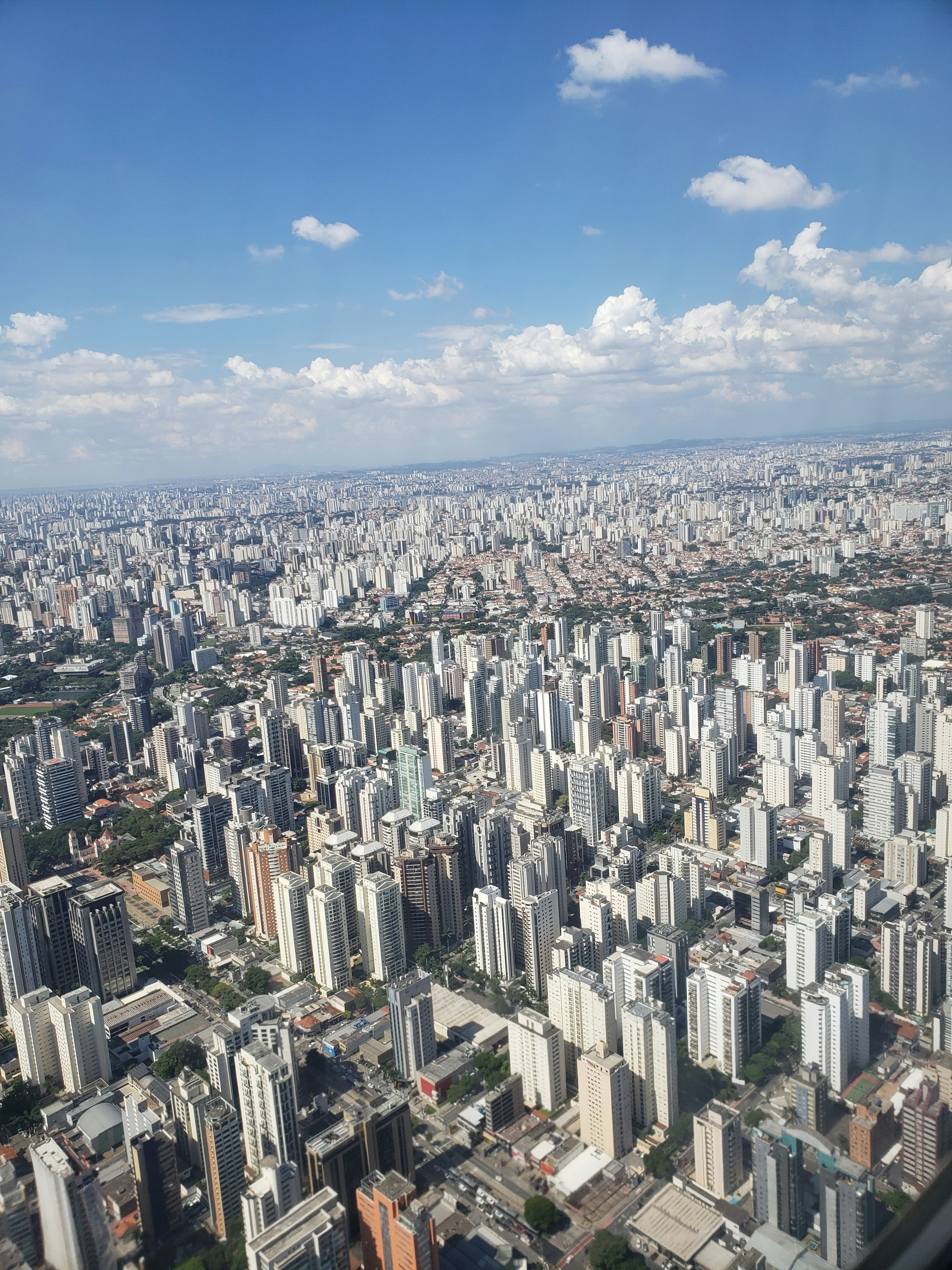 Aerial view of a sprawling cityscape, showcasing a dense arrangement of high-rise buildings and urban infrastructure under a bright blue sky with scattered clouds.