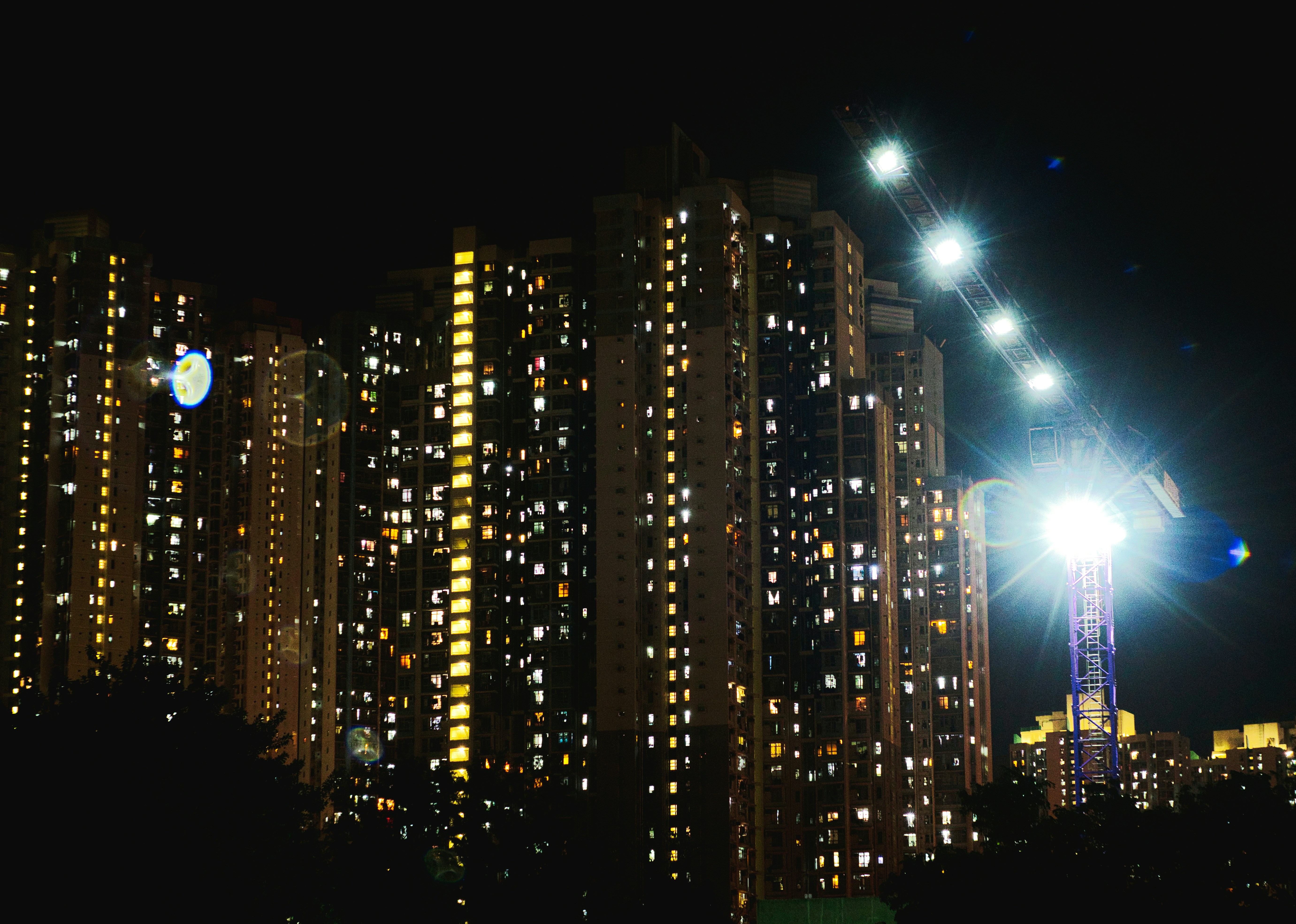Illuminated skyscrapers stand tall against the night sky, with a construction crane radiating bright light in the foreground.
