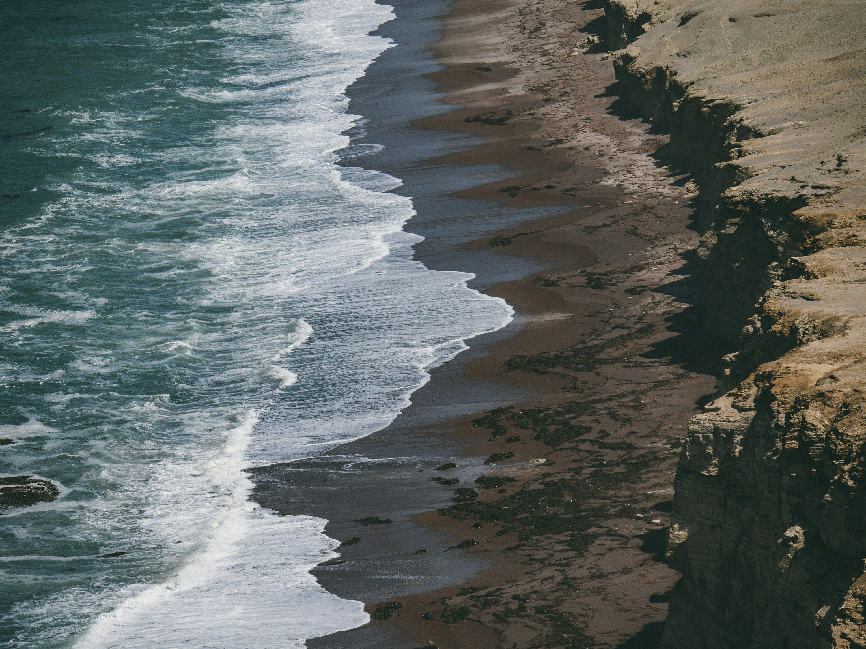 Waves gently lap against a rugged shoreline, revealing the contrast between dark sand and vibrant ocean blue.