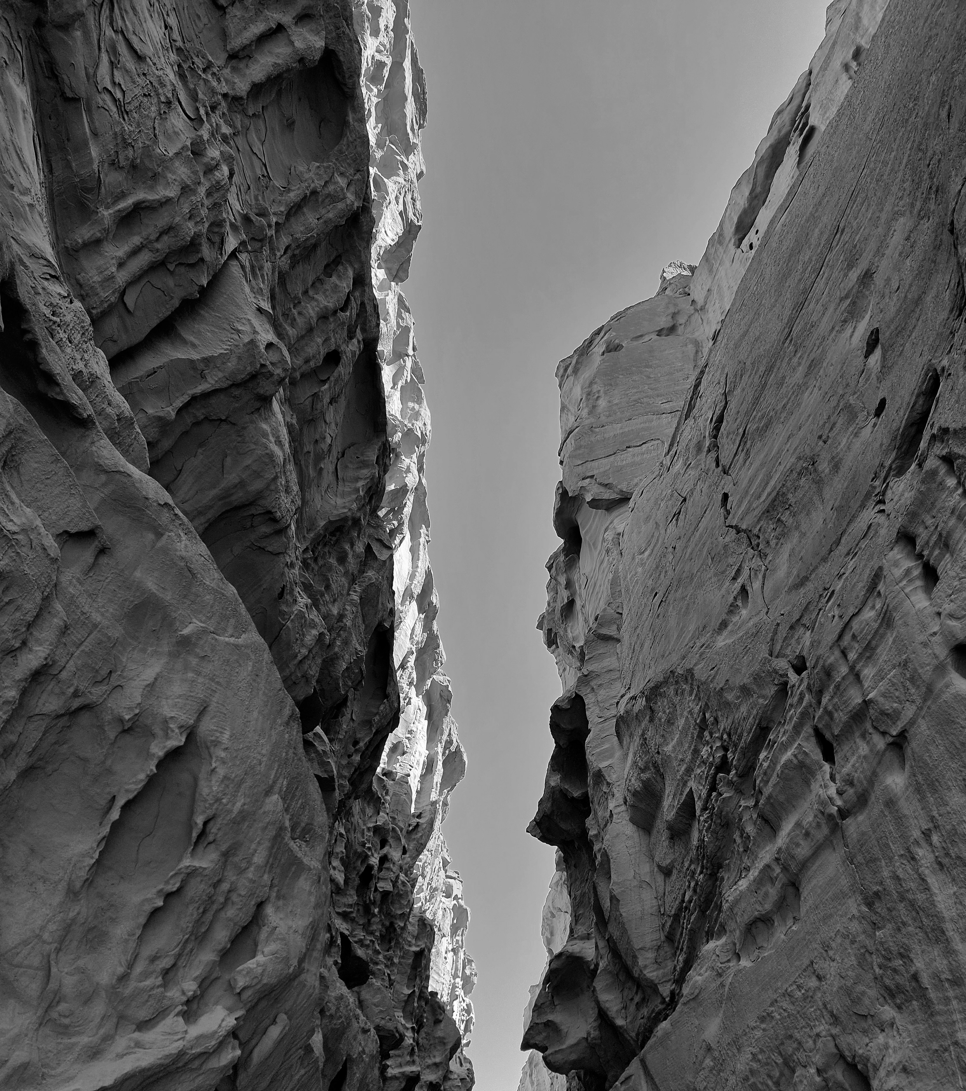 Vertical view of a narrow canyon with towering rock formations, showcasing intricate textures and shadows. The stark black and white contrast highlights the geological details.
