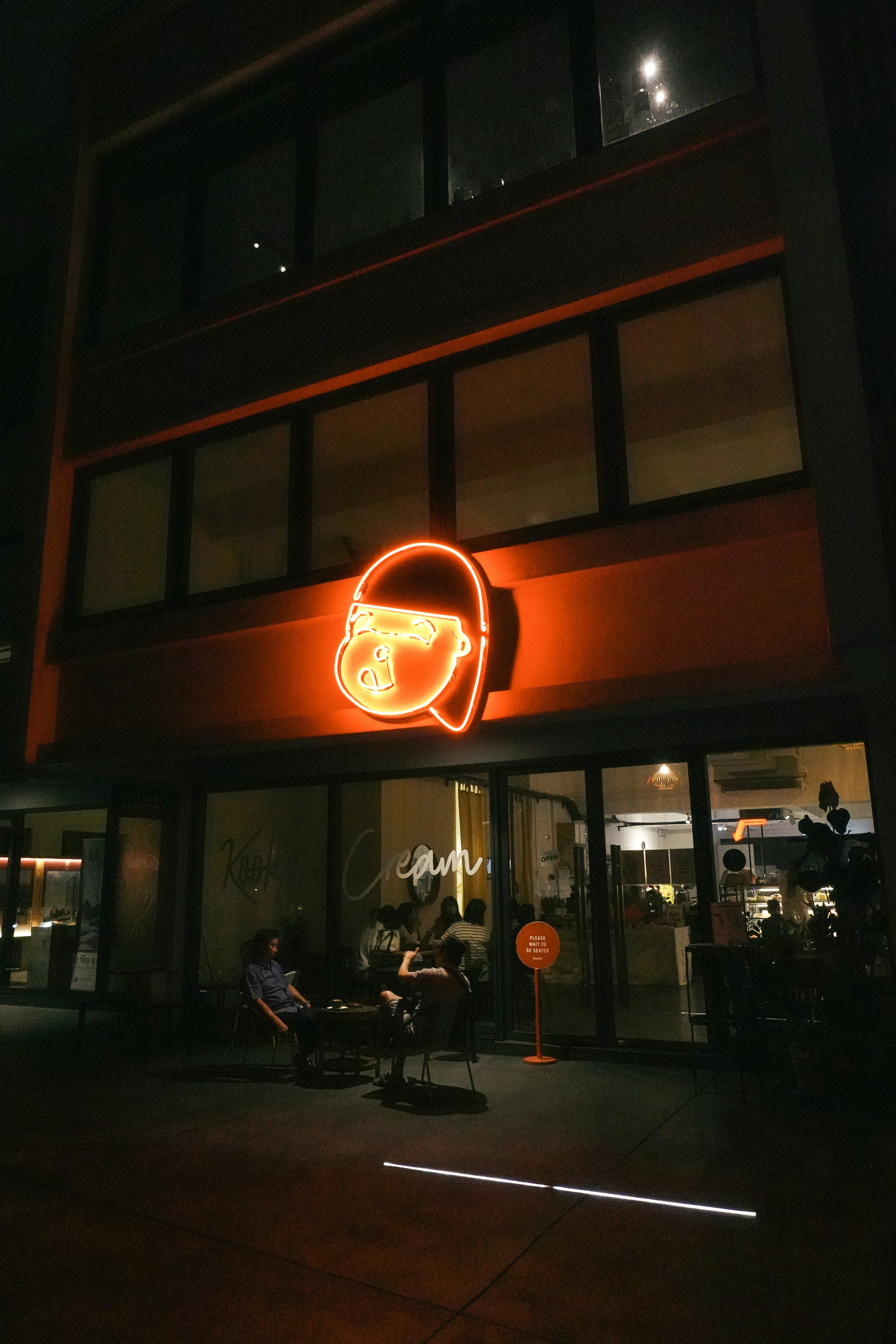 Neon sign featuring a stylized face illuminates a café entrance at night, with patrons seated outside enjoying the ambiance.