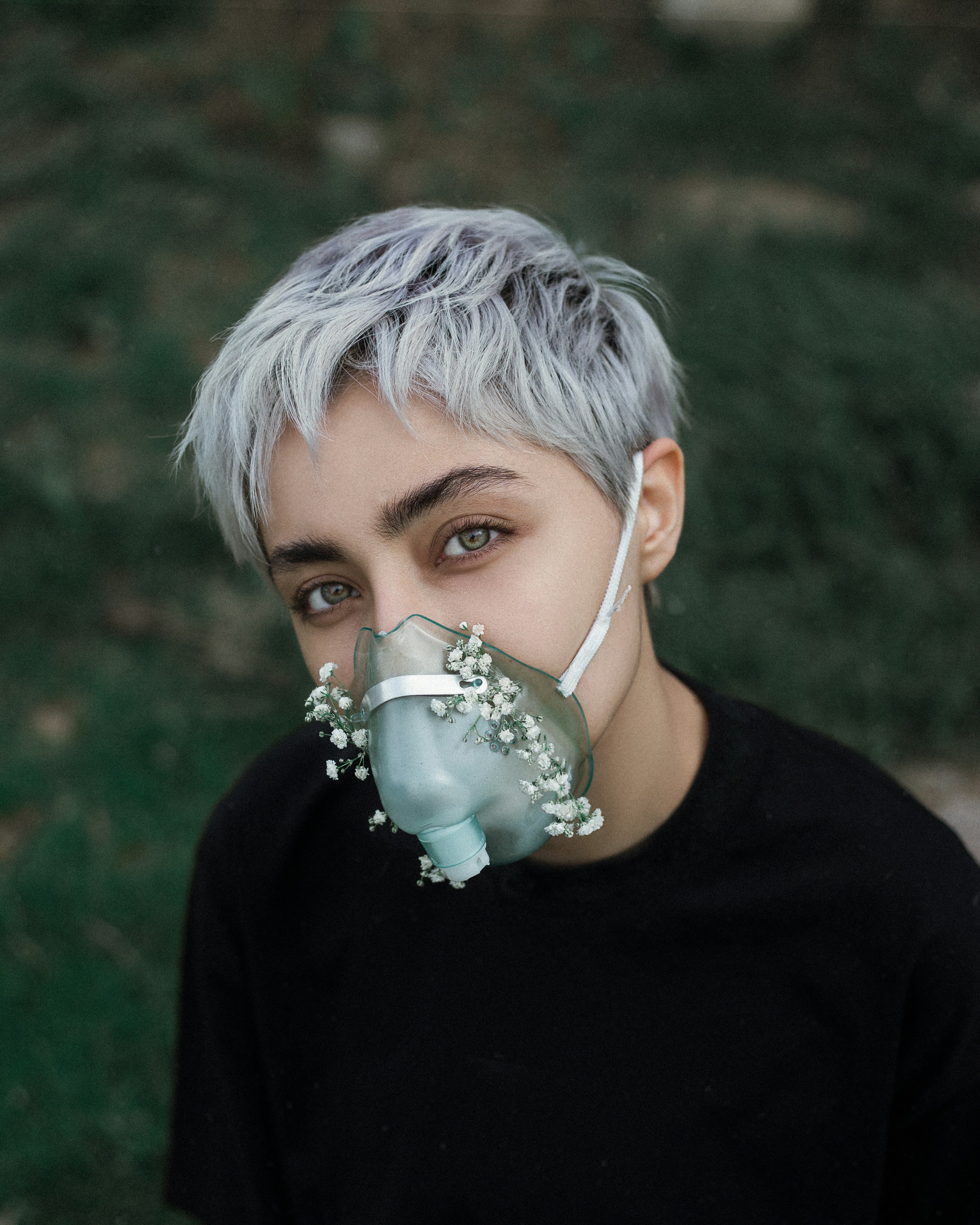 Individual with short, silver hair wearing a floral mask, gazing thoughtfully. The background features muted greens, enhancing the subject's presence.