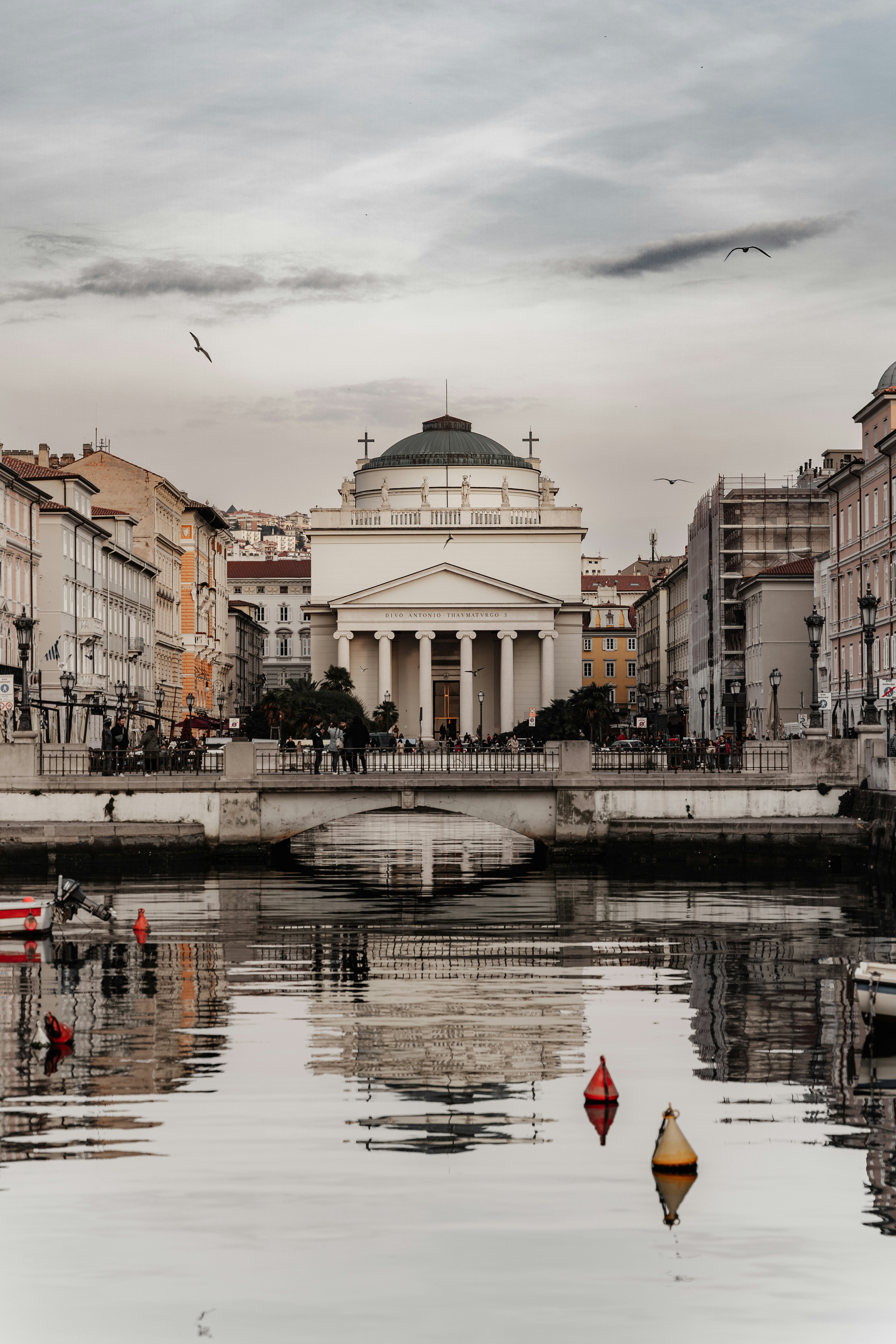 Neoclassical building reflecting in calm waters, framed by colorful facades and boats. The scene captures a serene urban landscape.