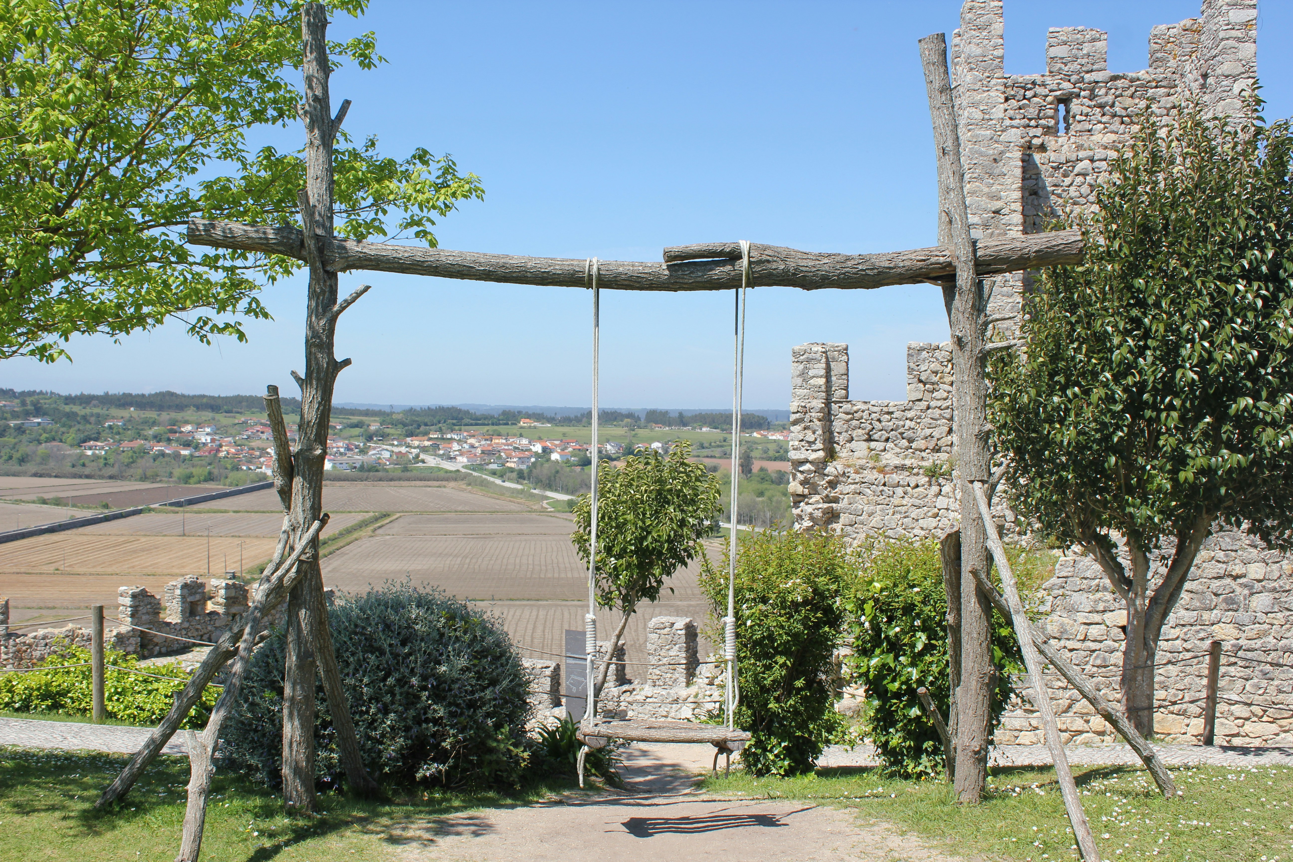 Rustic wooden swing set against a backdrop of a medieval stone castle and expansive countryside under a clear blue sky.