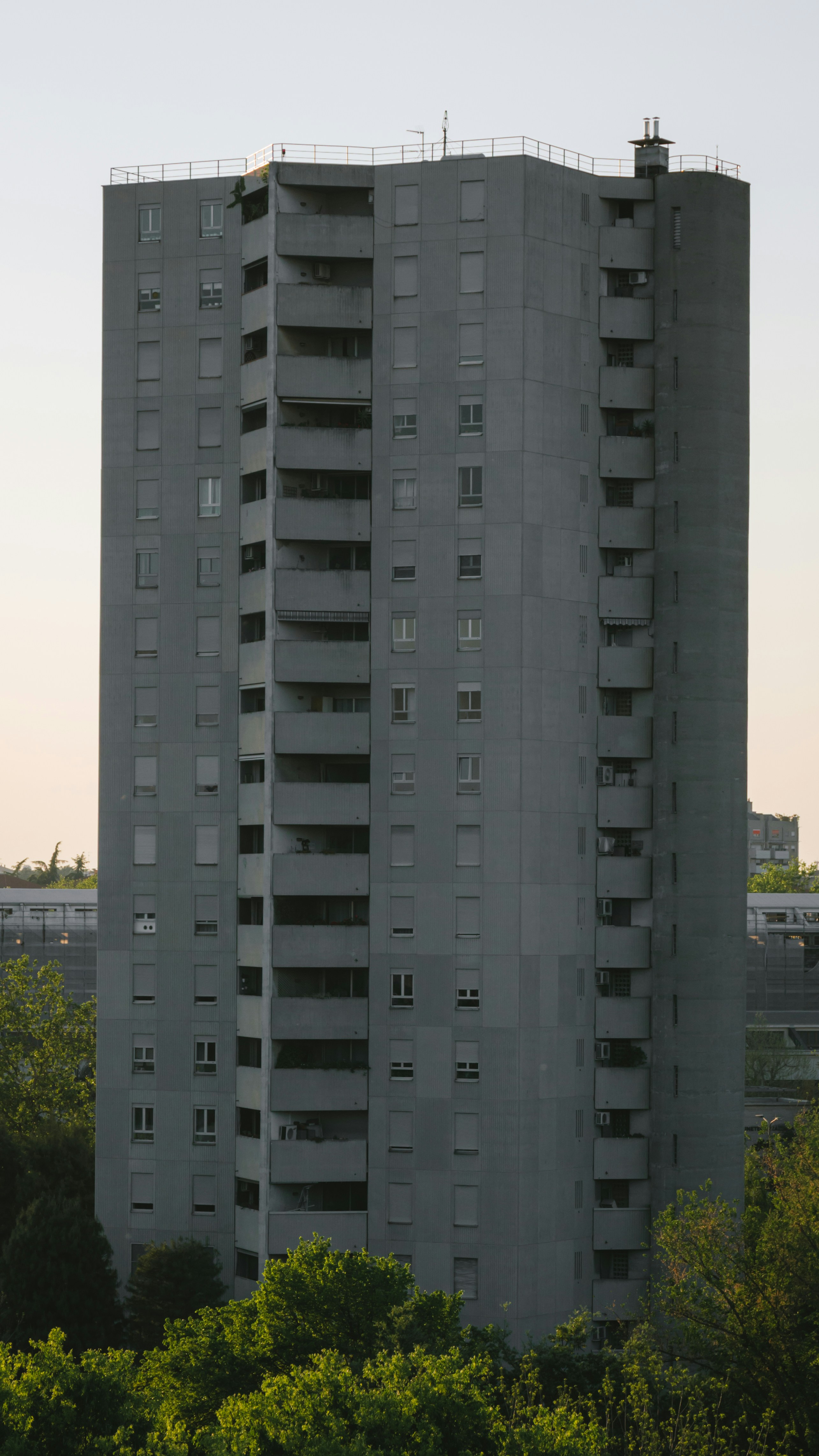 A tall, gray residential building stands prominently against a soft evening sky, surrounded by lush greenery at its base.