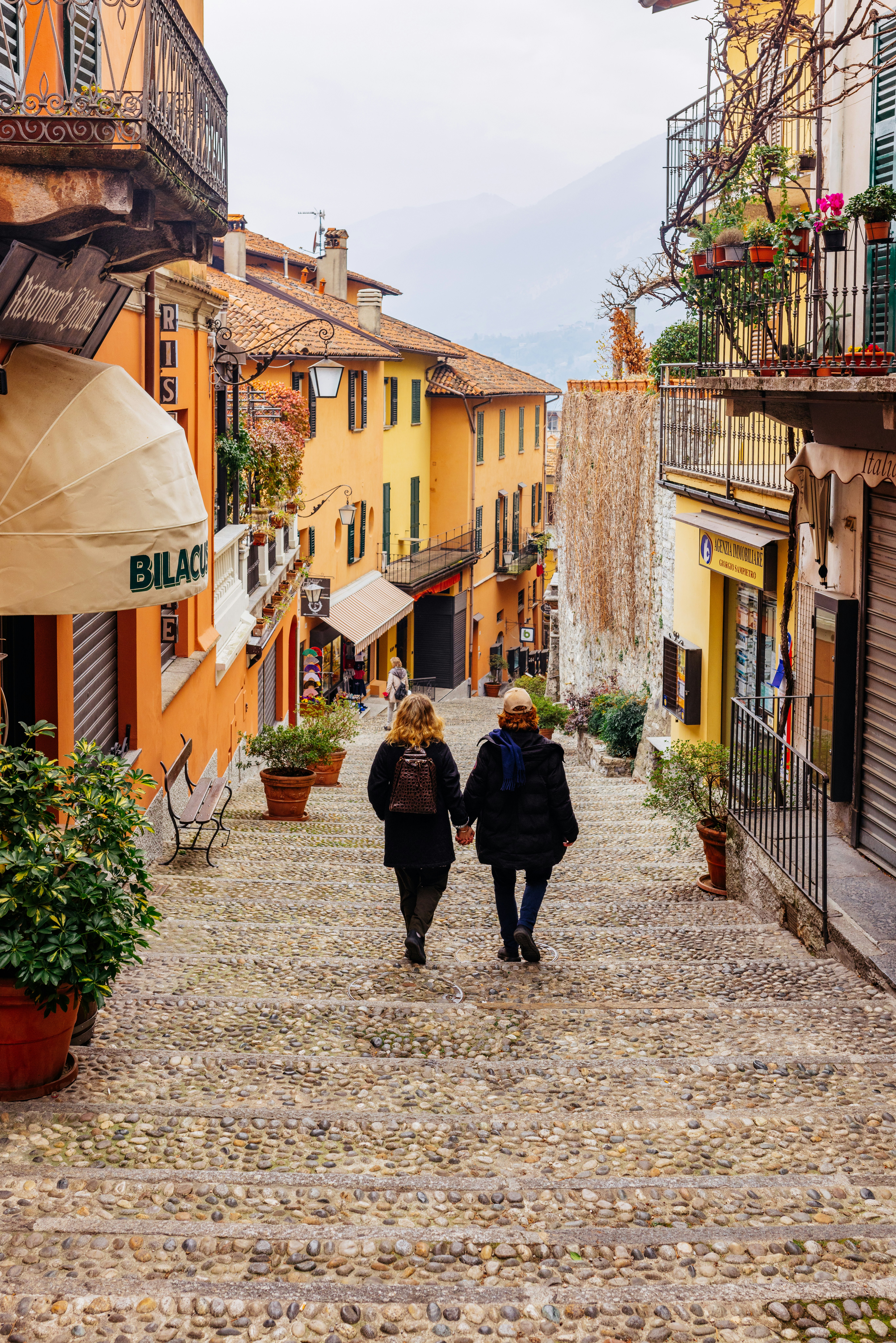 Two women stroll hand in hand down a cobblestone street lined with vibrant buildings and potted plants. The scene captures the charm of a quaint town.
