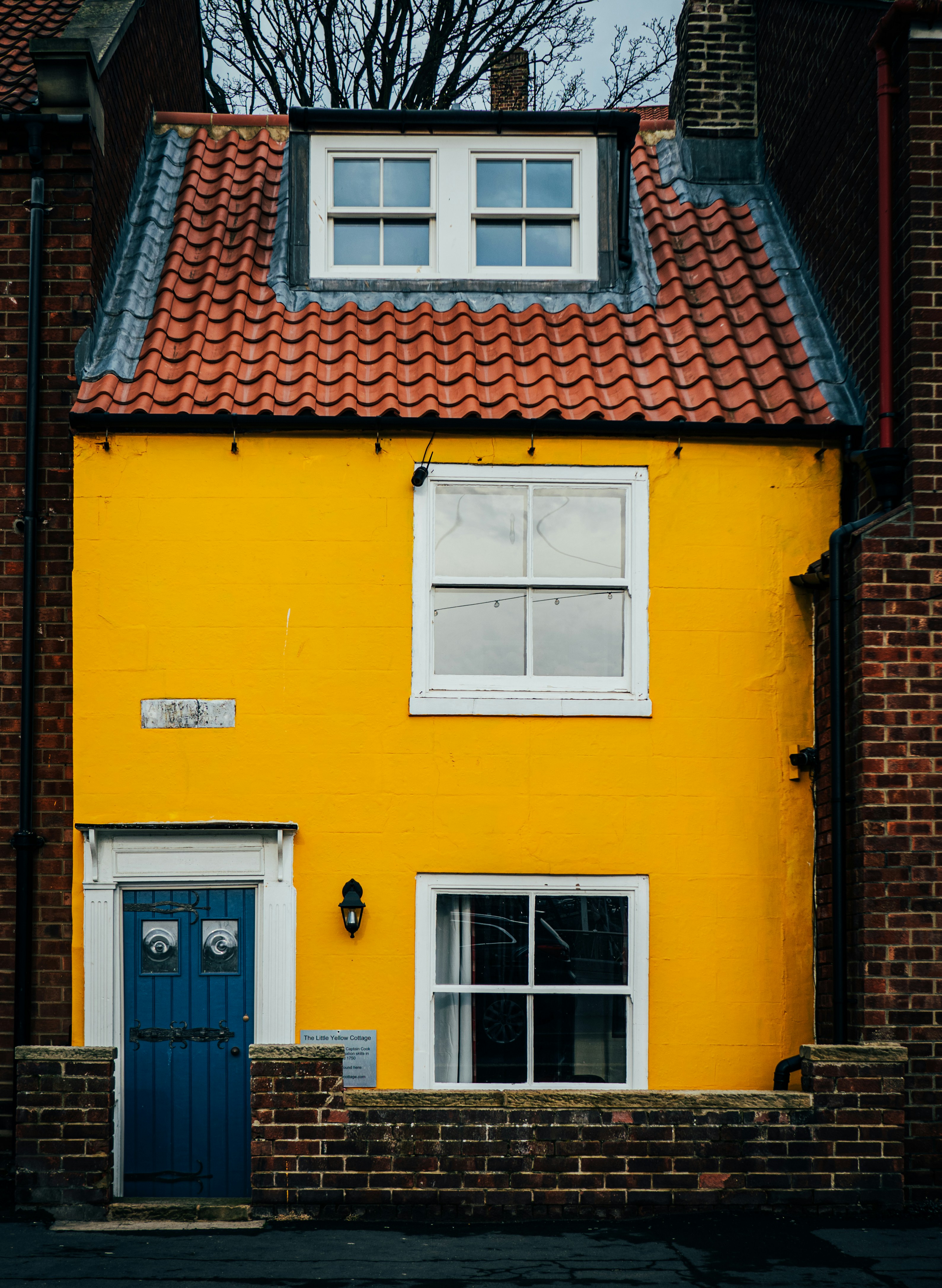 Vibrant yellow house with a blue door and charming windows, nestled between brick structures. A playful contrast of colors enhances the inviting character of this unique abode.