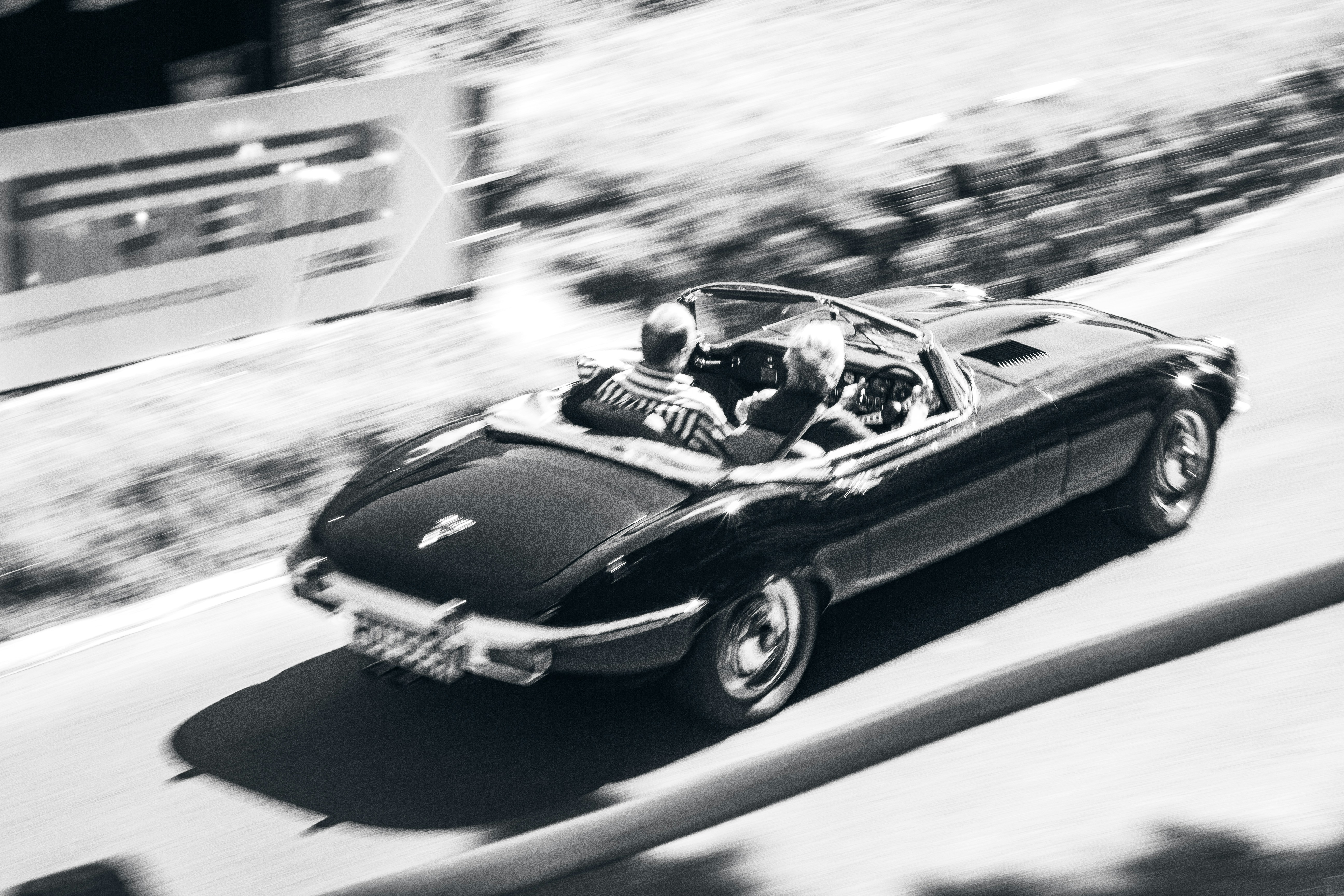 Black and white photograph of a vintage convertible car with two passengers driving on a sunny road.