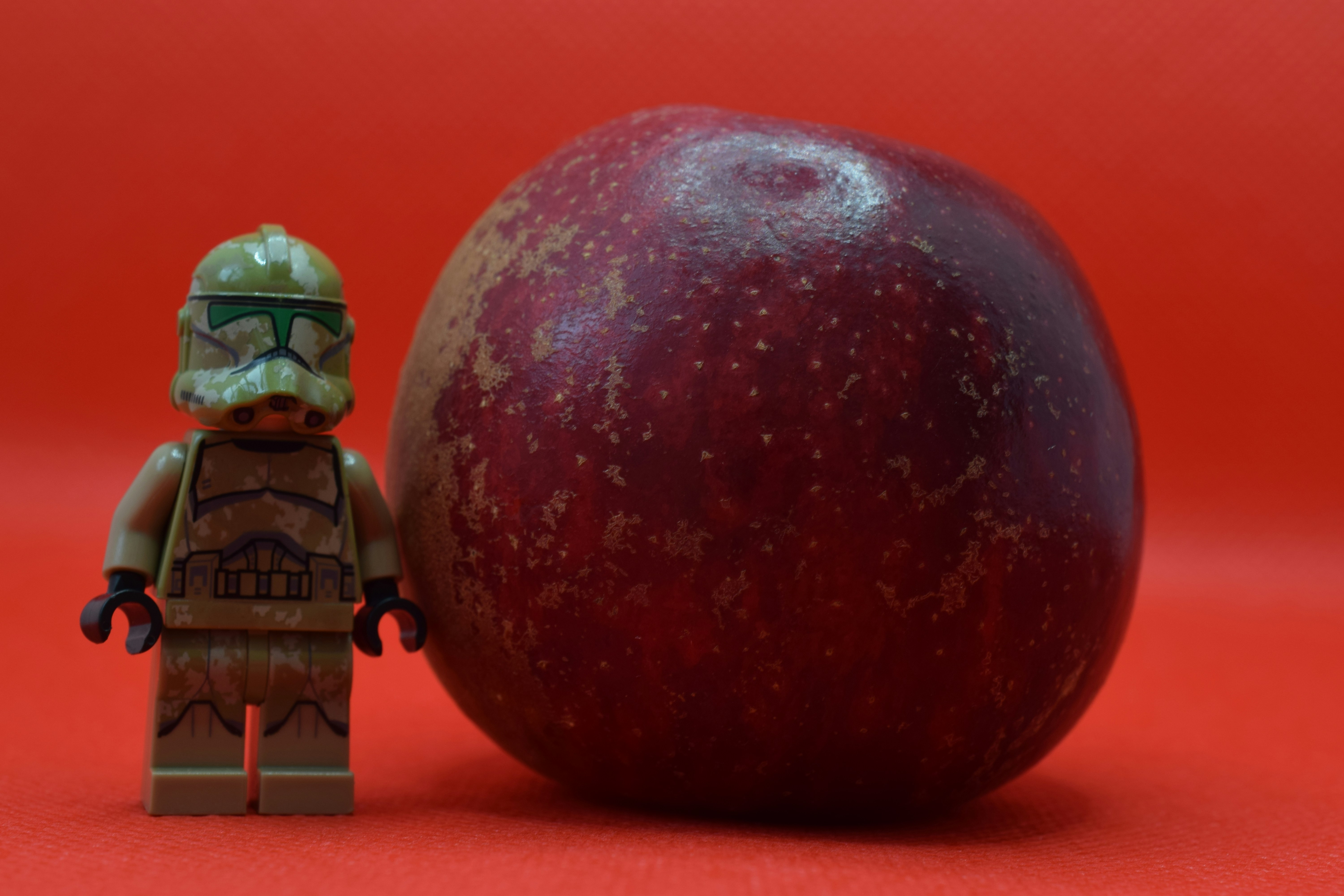 A LEGO stormtrooper stands beside a large red apple against a vibrant red background.