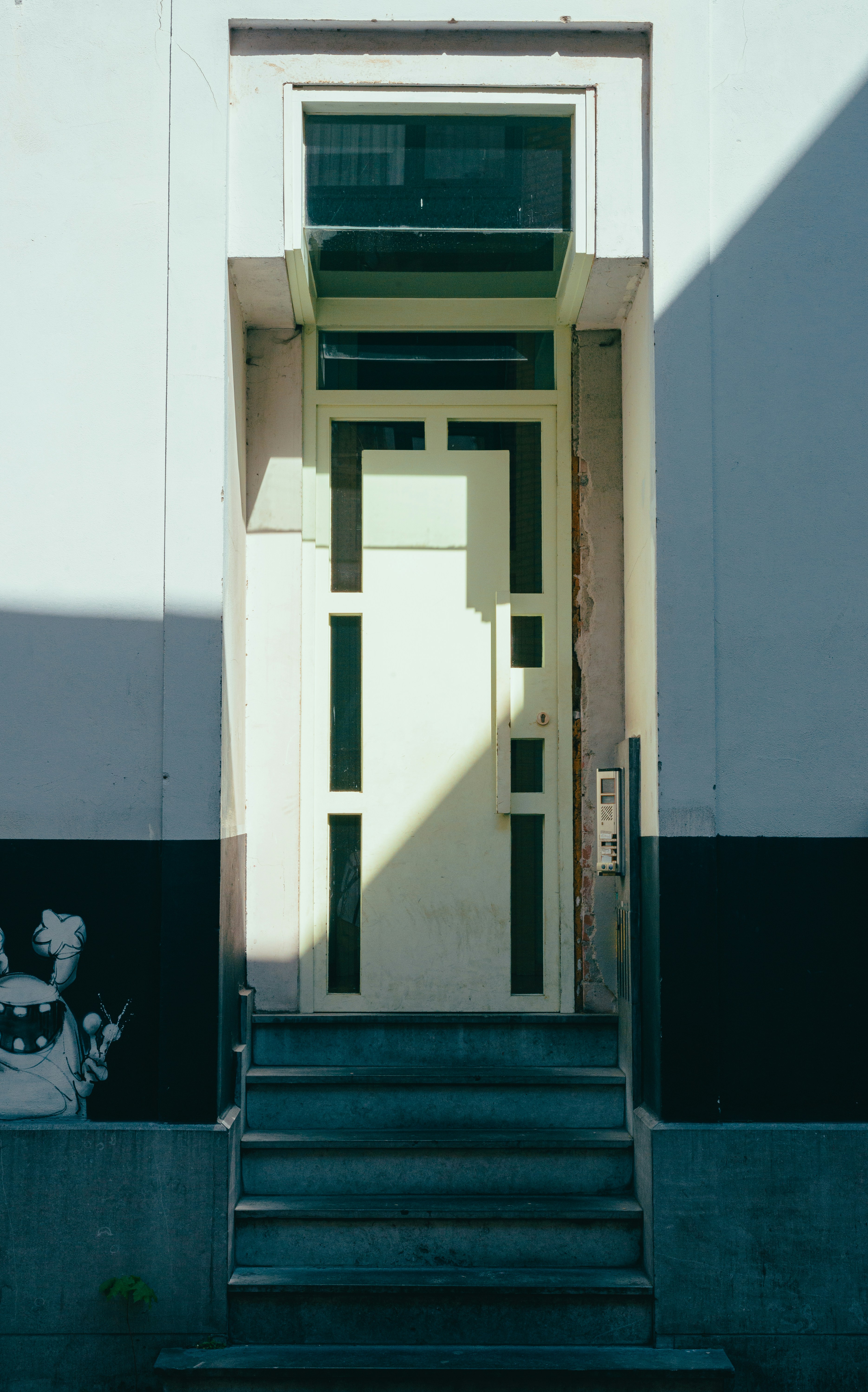 A vintage green door framed by a stark white and black wall, accompanied by playful street art on the left. The sunlight casts a dramatic shadow, enhancing the architectural features.
