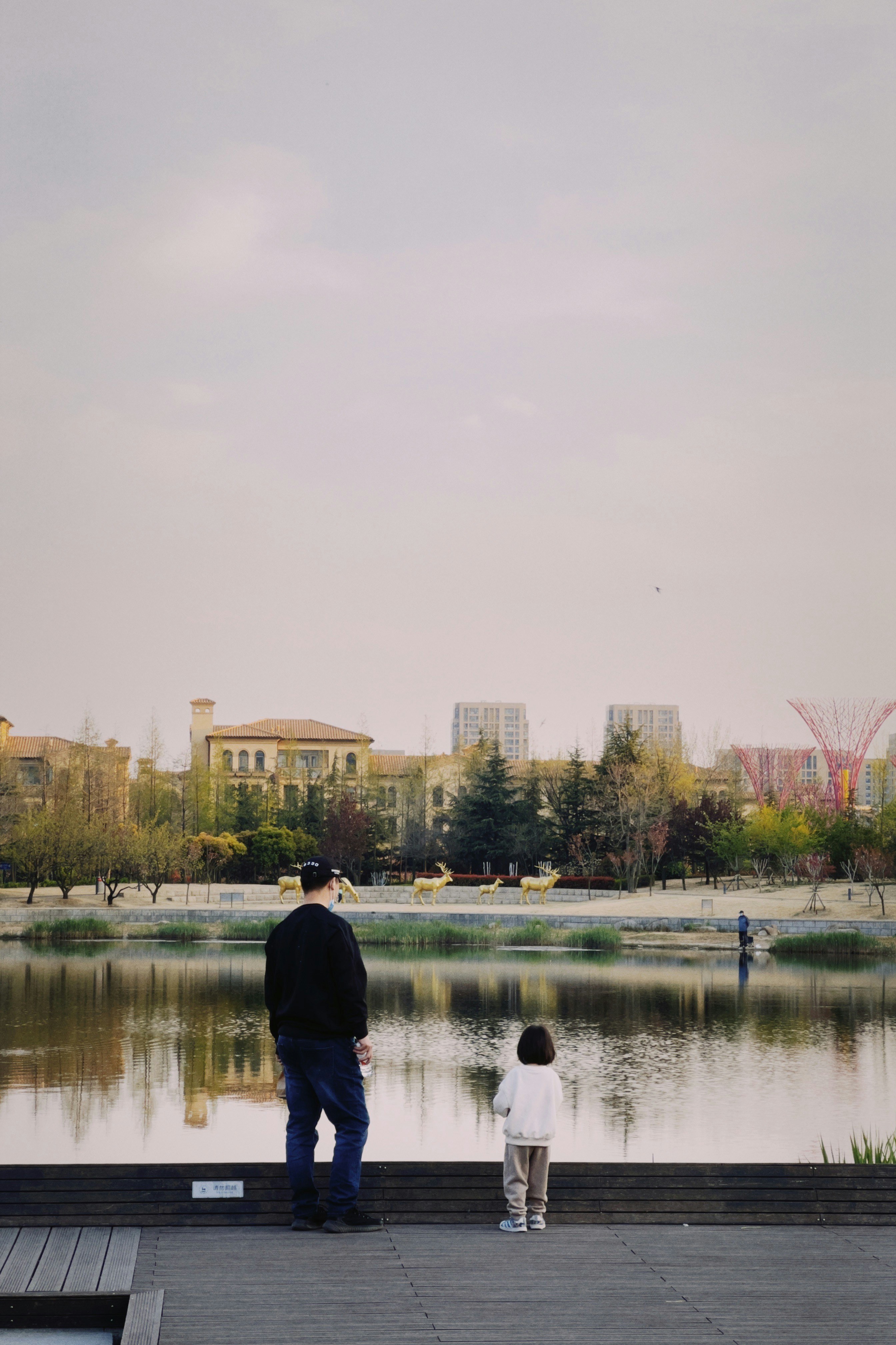 Man and child stand on a wooden deck overlooking a tranquil lake with distant buildings and sculptures.