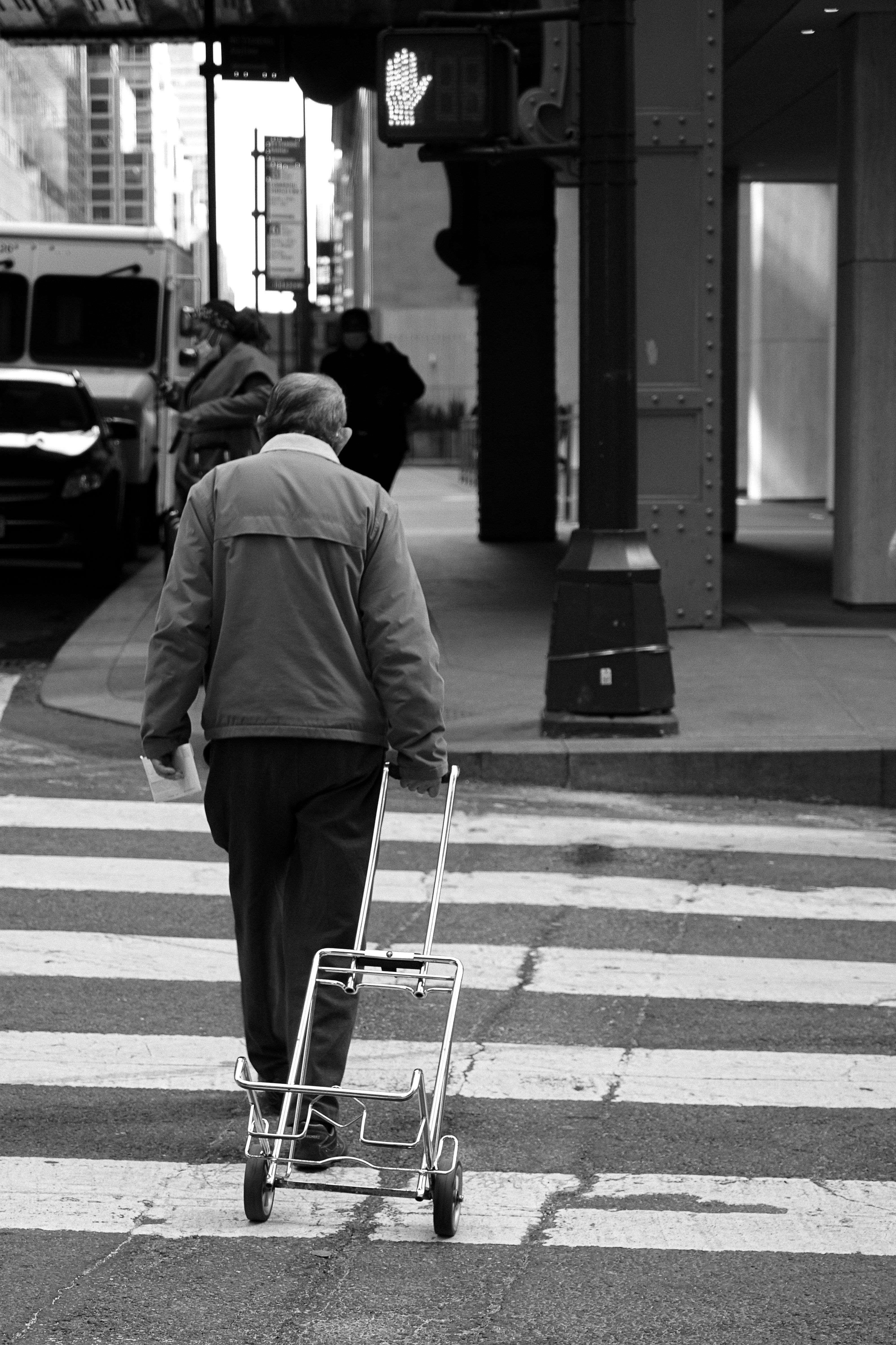 An elderly man navigates a city crosswalk, pulling a shopping cart, framed by urban architecture. The scene captures the essence of daily life in monochrome.