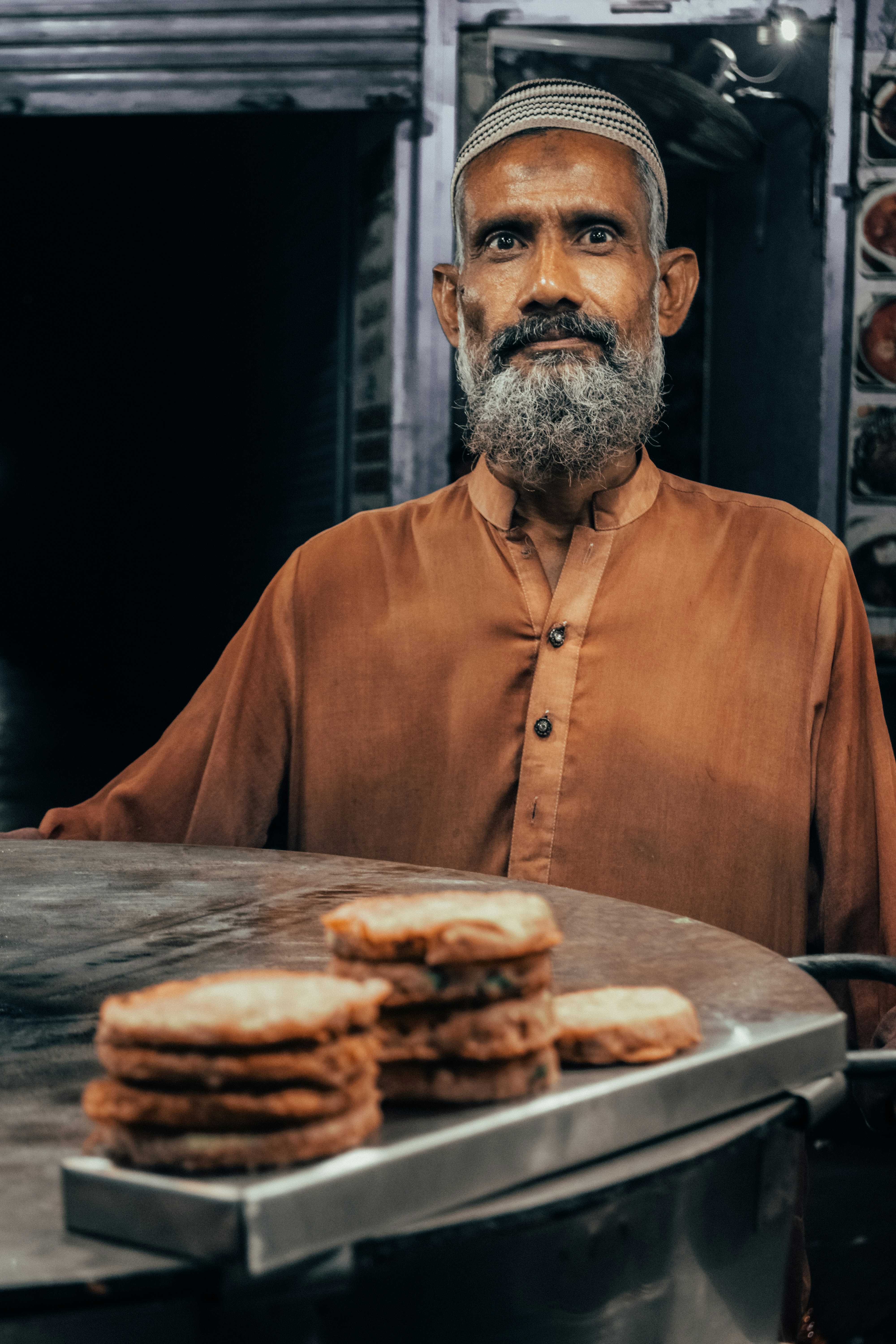 A seasoned vendor stands behind a metallic counter, showcasing freshly made snacks, with a focus on his expressive demeanor and traditional attire.