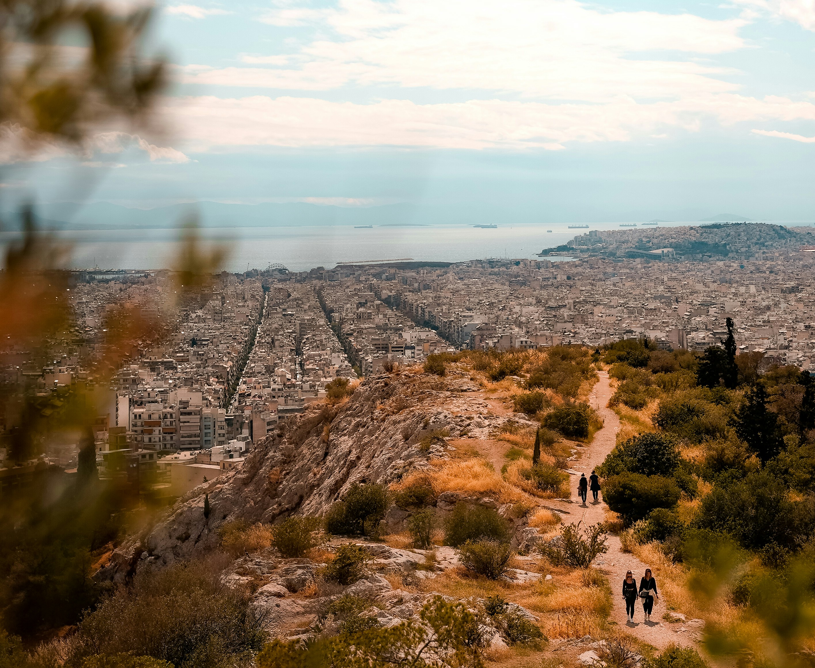A scenic view overlooking a sprawling cityscape with a winding path and hikers, framed by lush greenery. The horizon meets the sea in the distance.