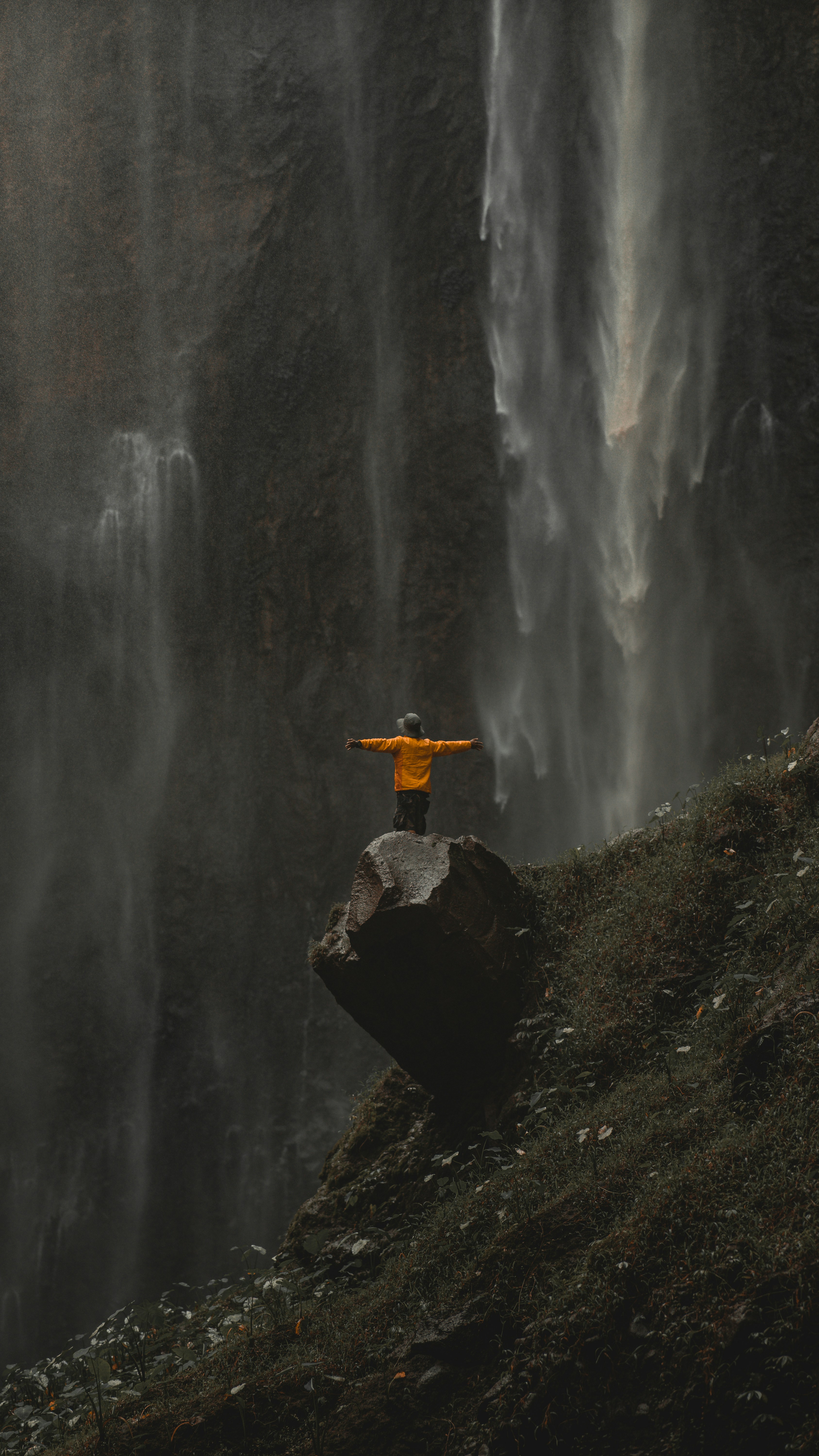 Person in a bright yellow jacket stands triumphantly on a rock, surrounded by cascading waterfalls in a dramatic landscape.